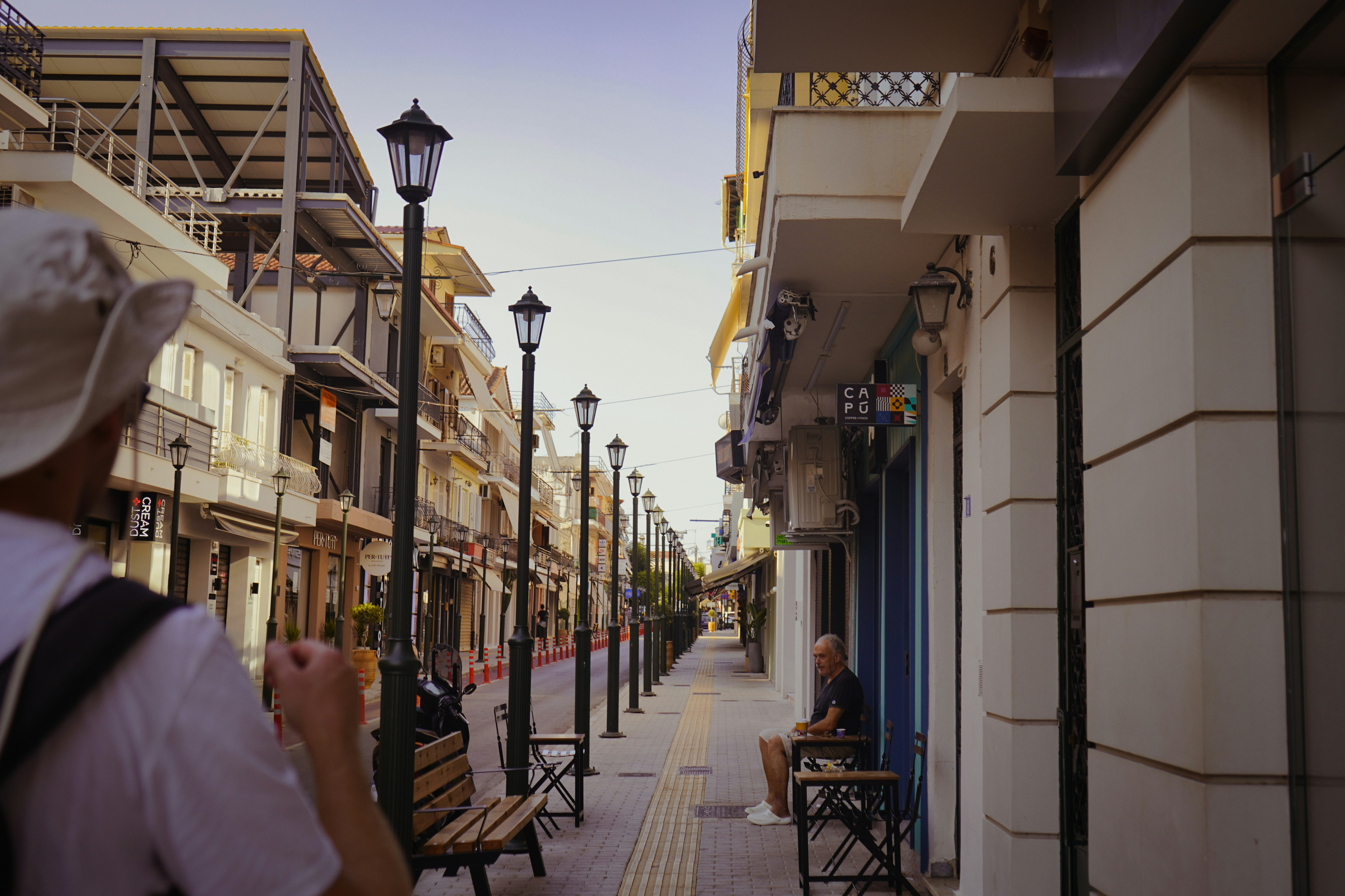 Person with a hat walking down a narrow, sunlit street lined with lampposts and buildings.