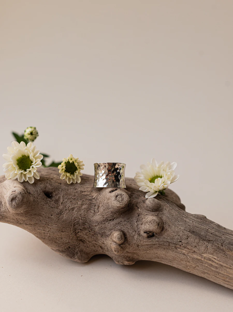 A silver ring sits on a piece of weathered driftwood accented by small white flowers against a soft neutral background.