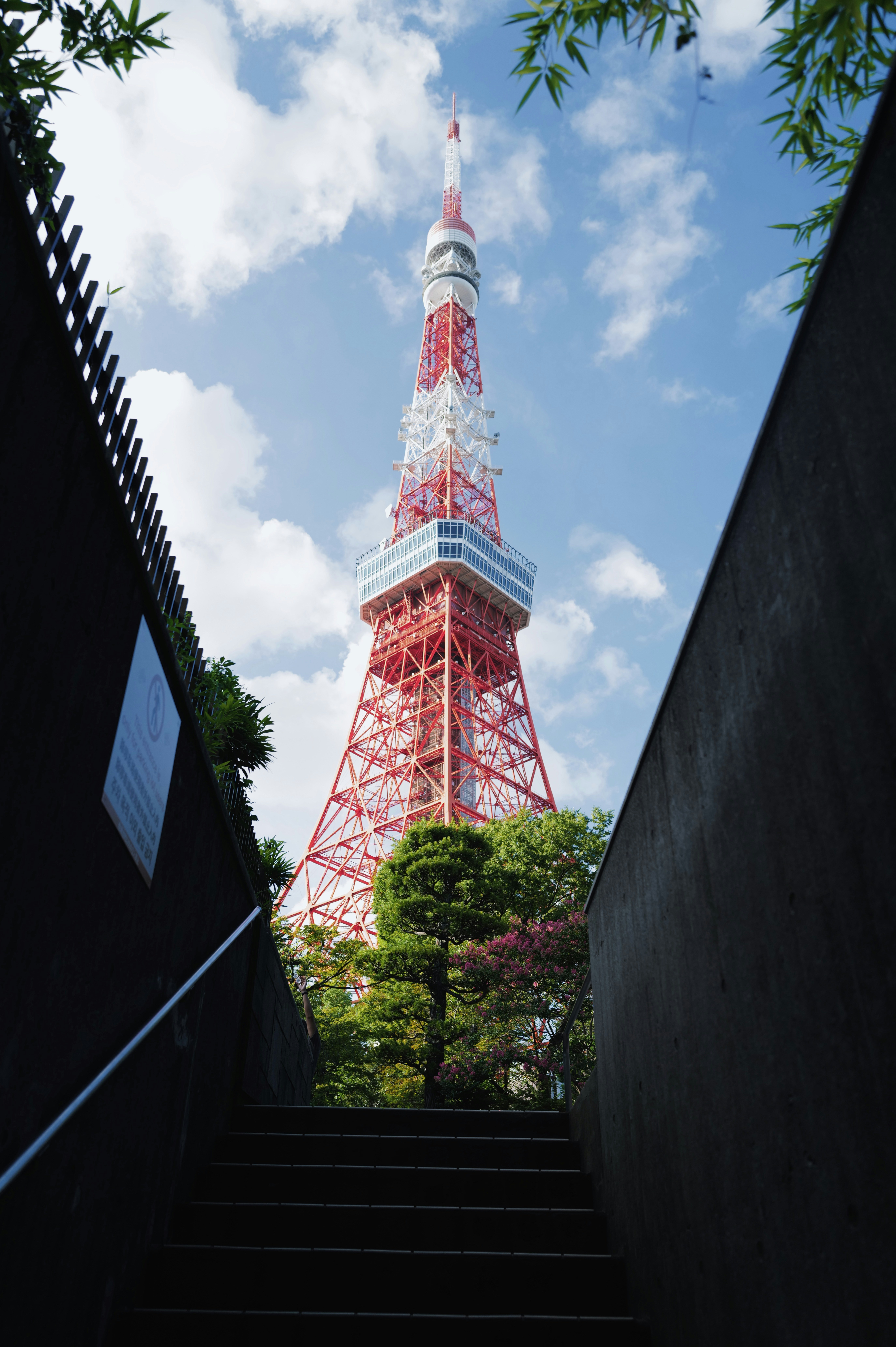 Tokyo Tower rises through a framed alley, its red lattice set against a bright blue sky. The composition emphasizes the contrast between the shadowed foreground and the tower's intricate structure.