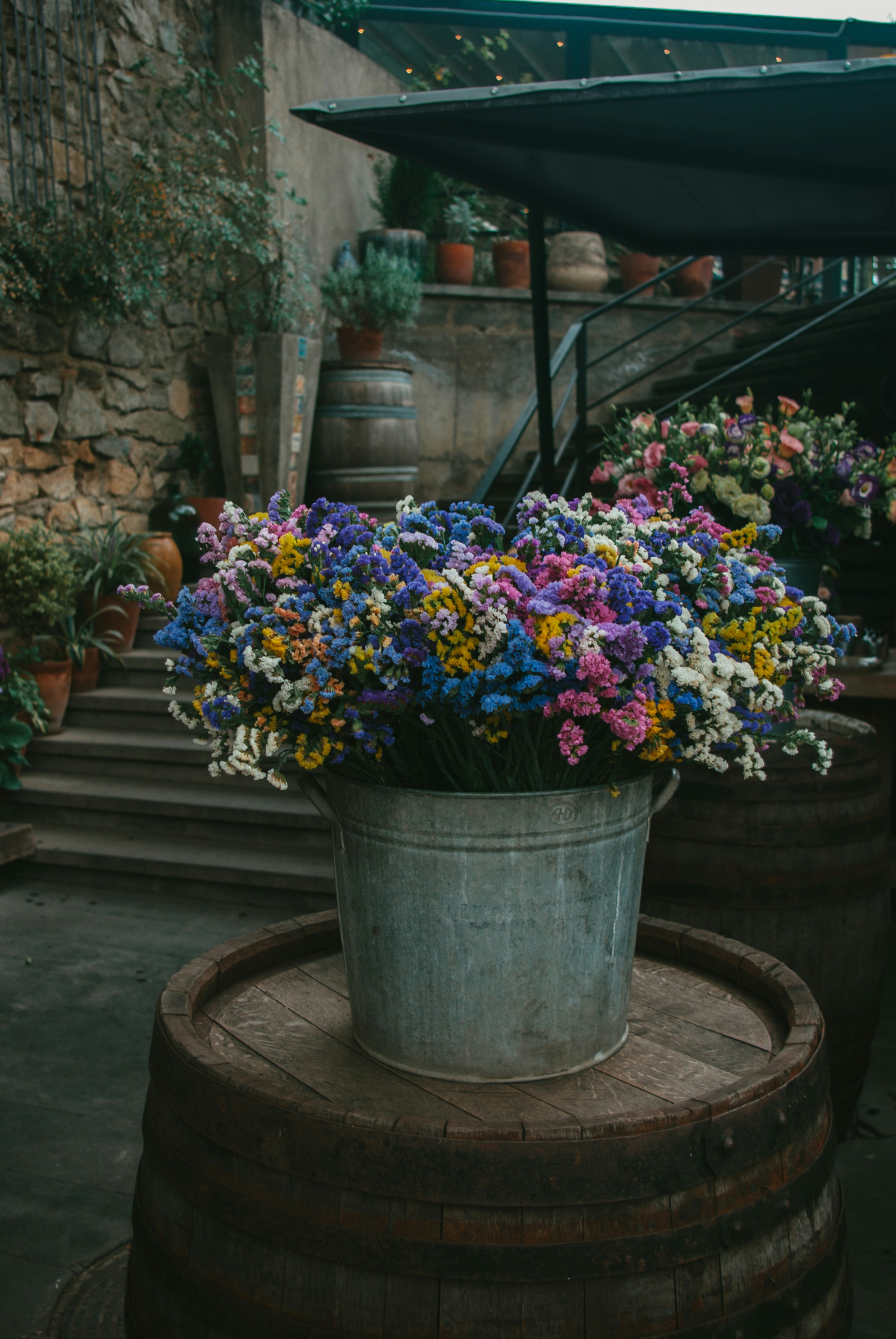 A metal pail brimming with a multicolored bouquet sits atop a wooden barrel in a rustic courtyard, with stone stairs and potted plants in the background.