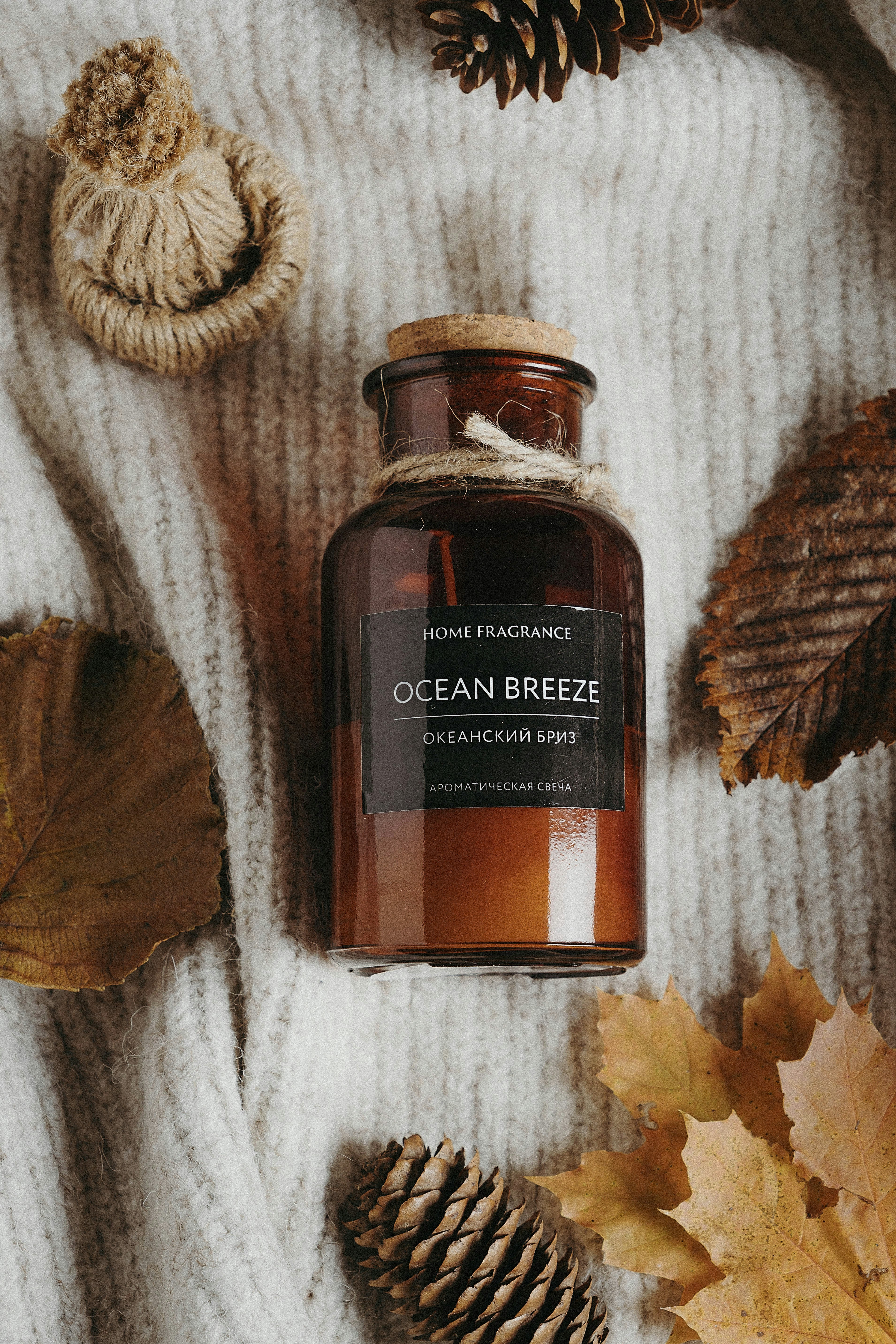 Amber glass candle jar labeled 'Ocean Breeze' surrounded by autumn leaves and pine cones on a textured knit background.