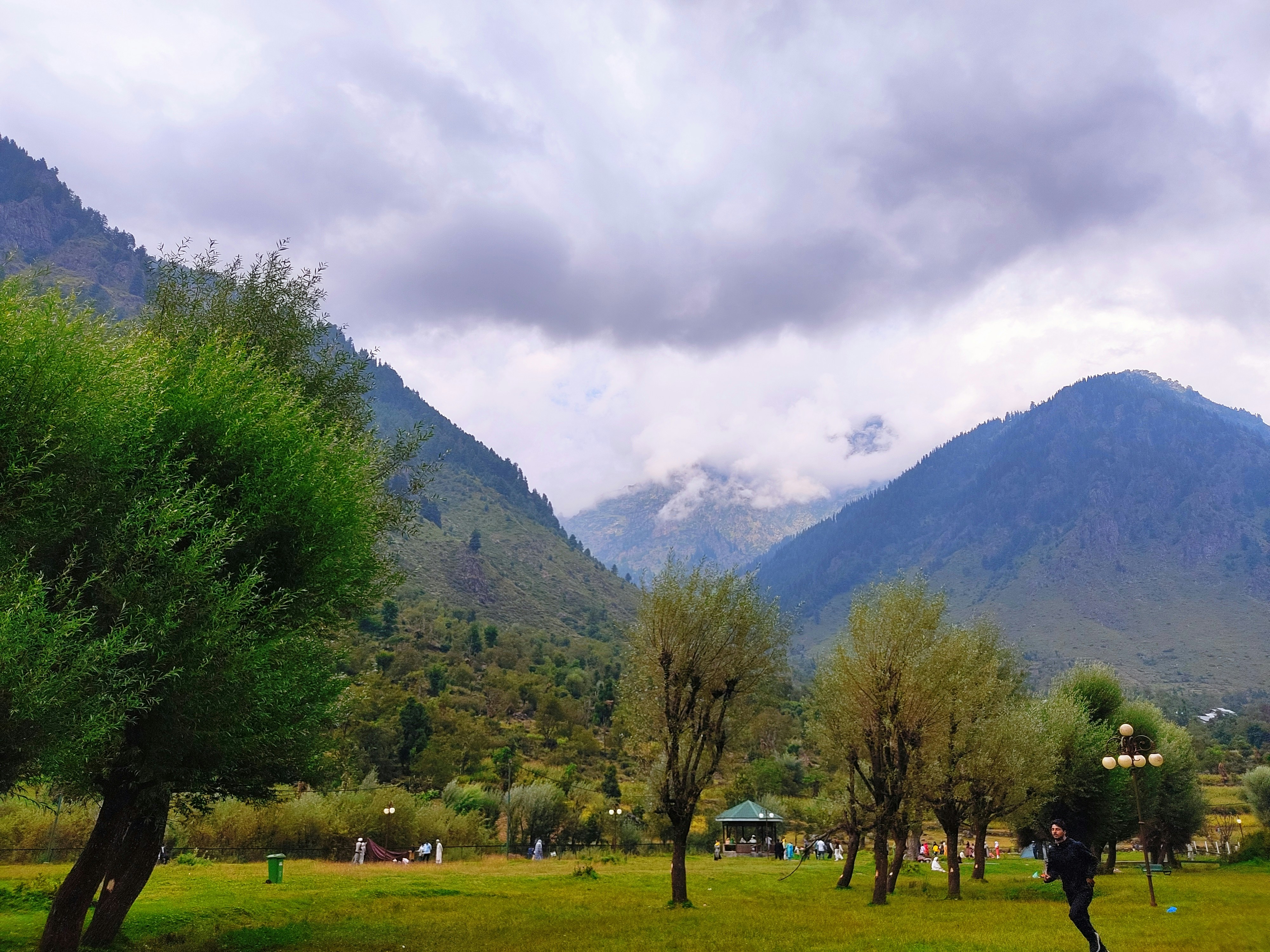 Landscape photograph of a park with trees, a small gazebo, and distant misty mountains under a cloudy sky.