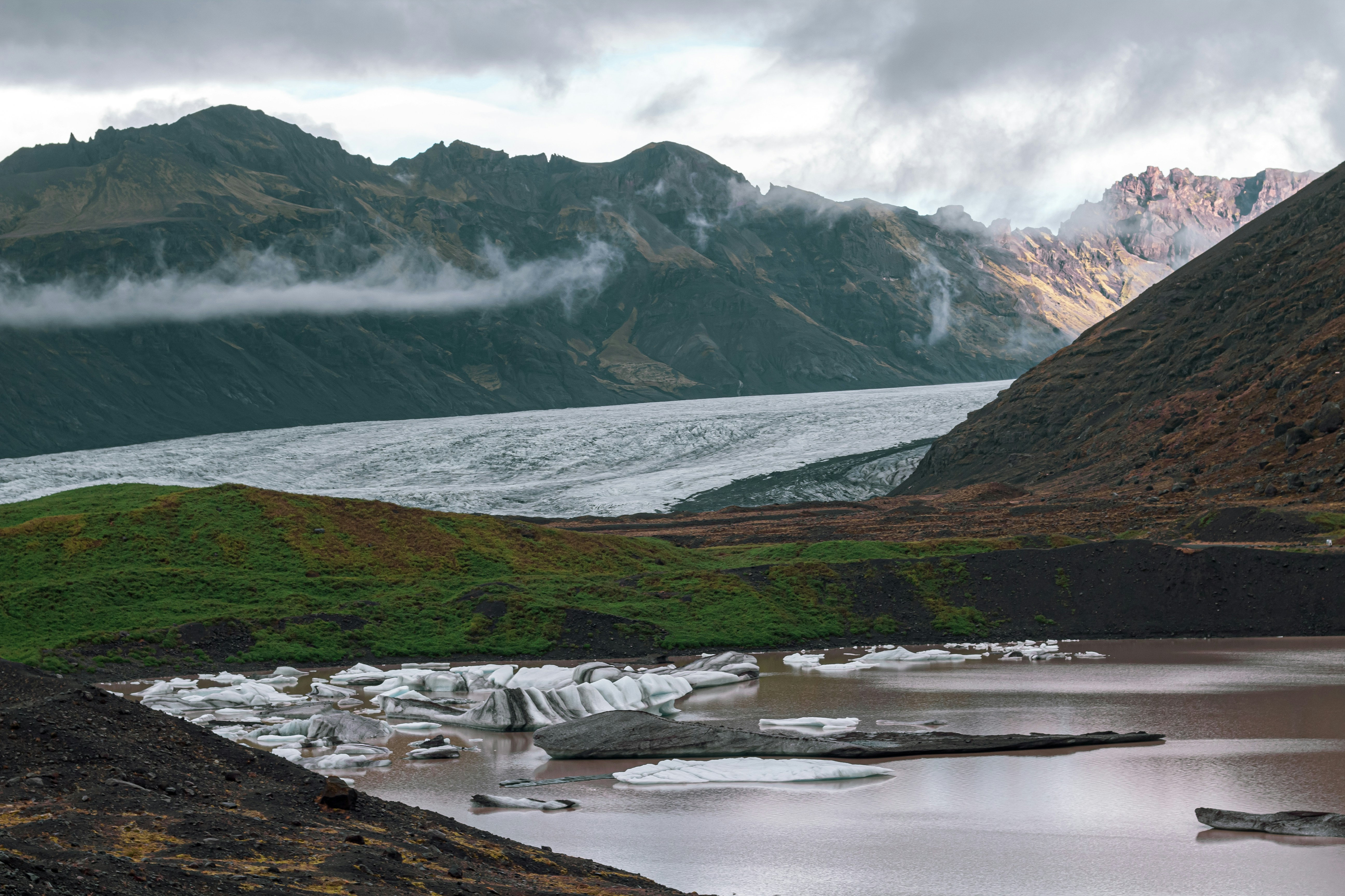 A tranquil scene showcasing a glacial lake surrounded by rugged mountains, with icebergs floating on the water. The lush green hills contrast beautifully with the stark ice and rocky terrain.