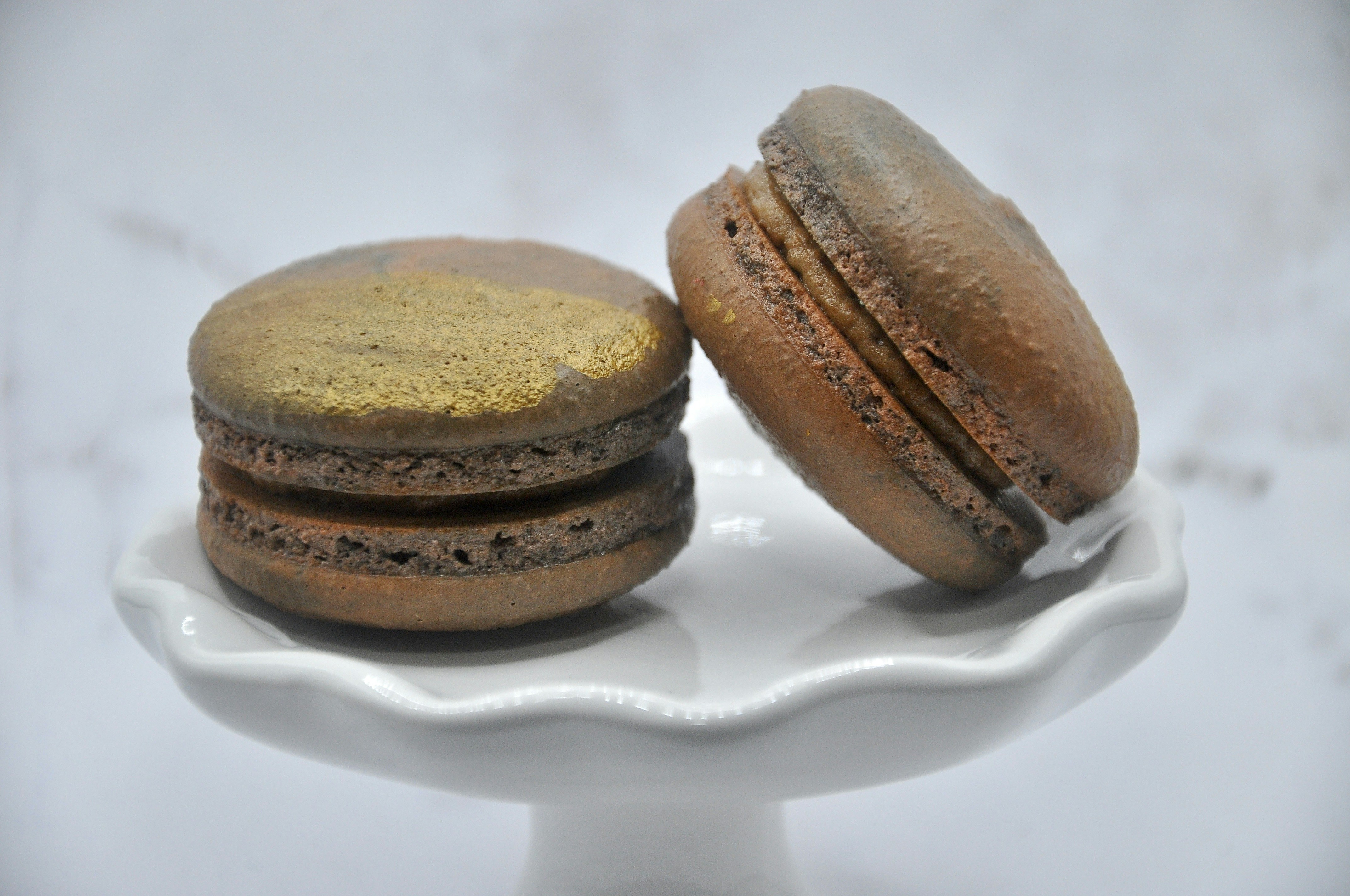 Two Cocoa Macarons Rest on a White Pedestal Plate Under Soft Studio Light