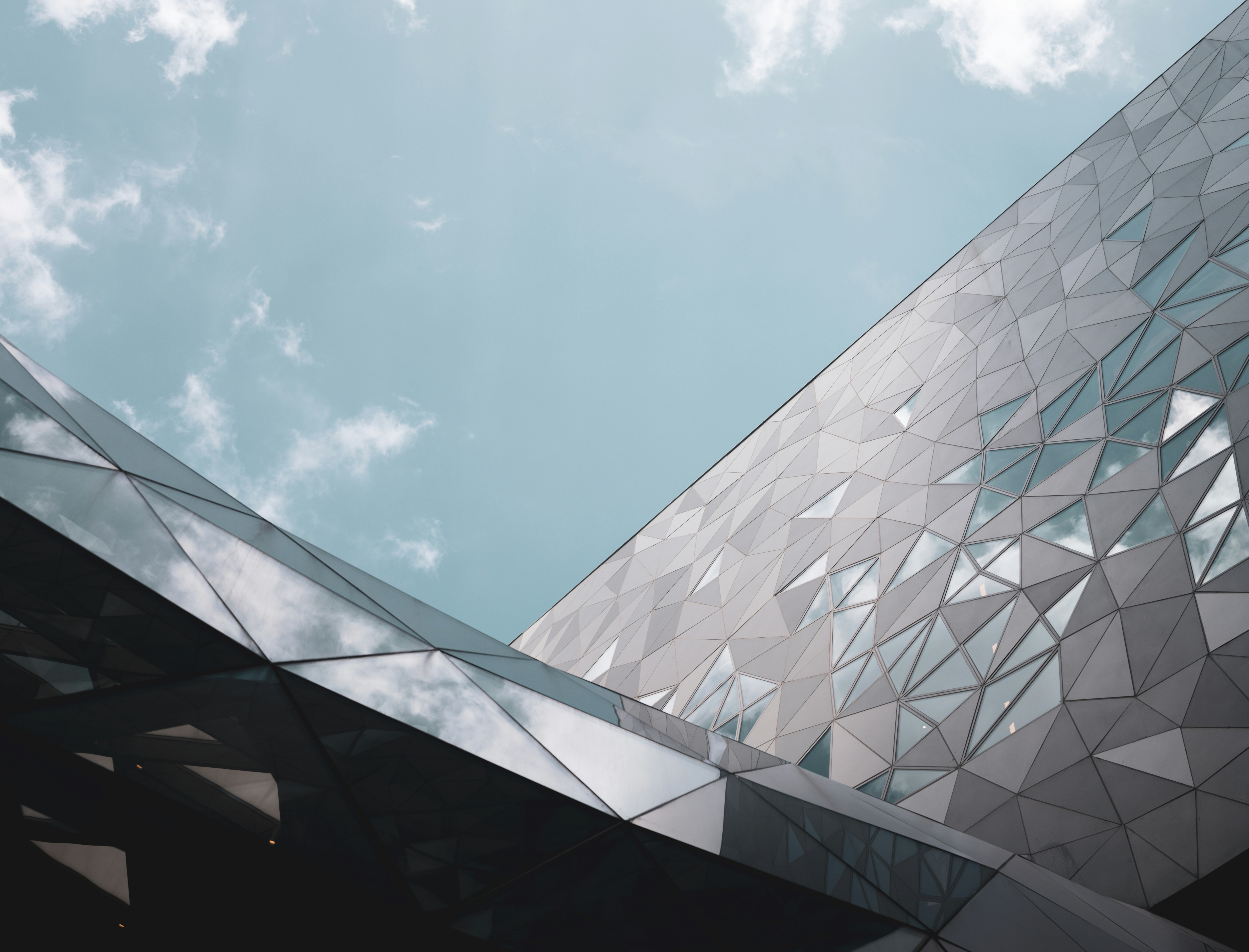 Architectural photograph of angular metal-clad surfaces converging along diagonal planes against a pale blue sky. The composition emphasizes geometry, reflections, and the interplay of light on the facets.