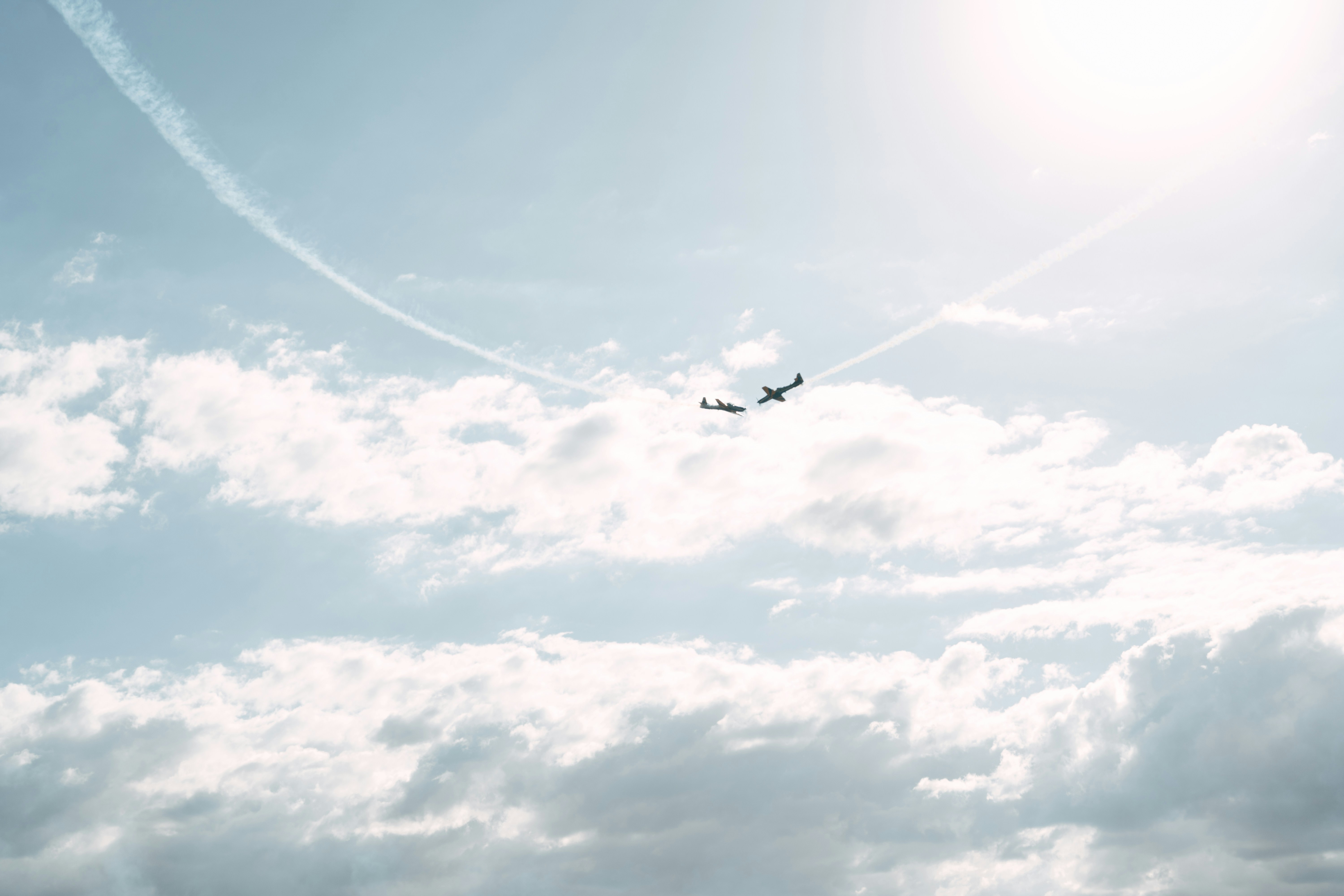 Aircraft performing aerobatic maneuvers with contrails against a bright sky filled with scattered clouds.