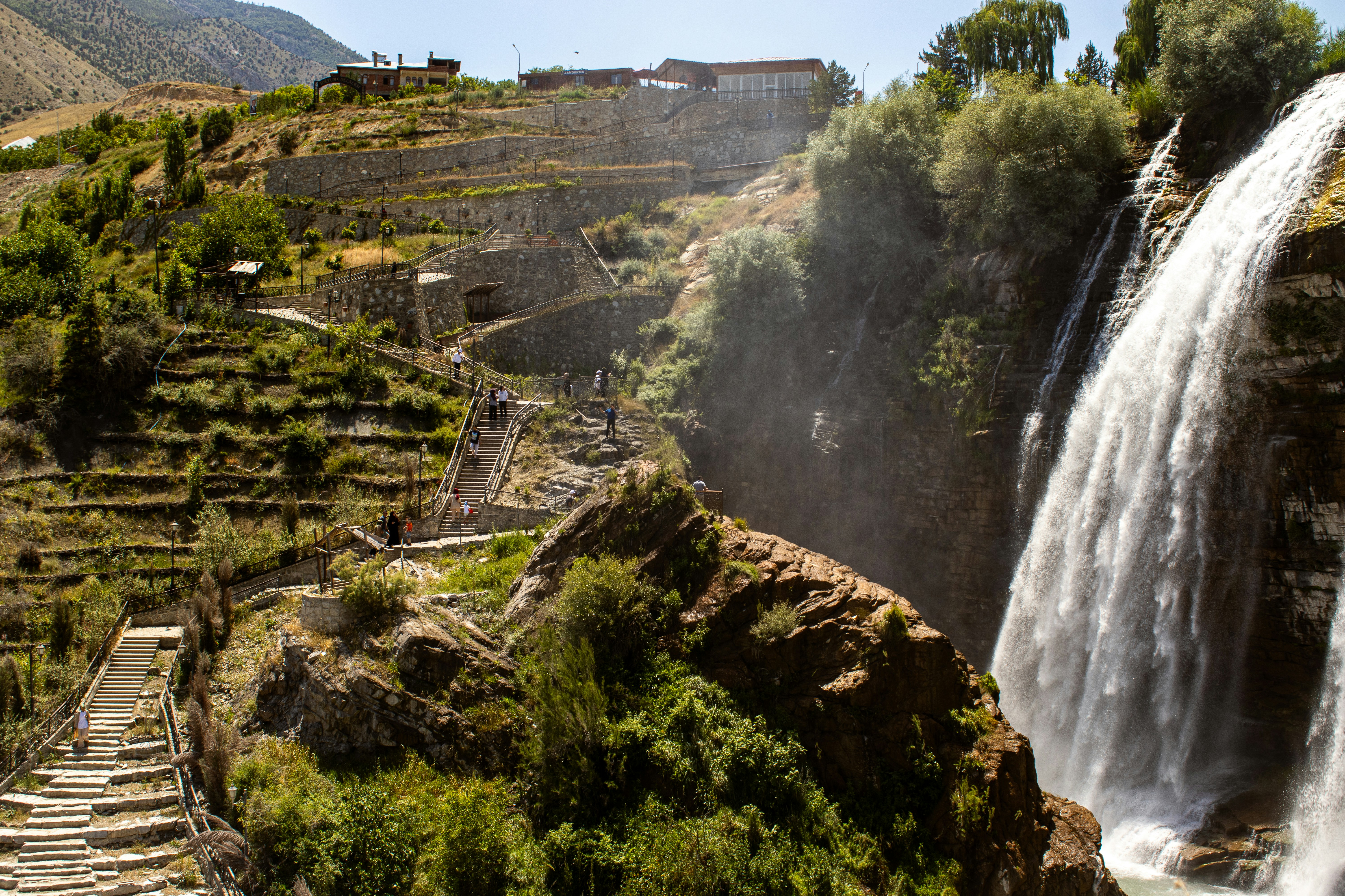 A waterfall cascades down rocky cliffs with terraced pathways and lush greenery surrounding it under a clear sky.