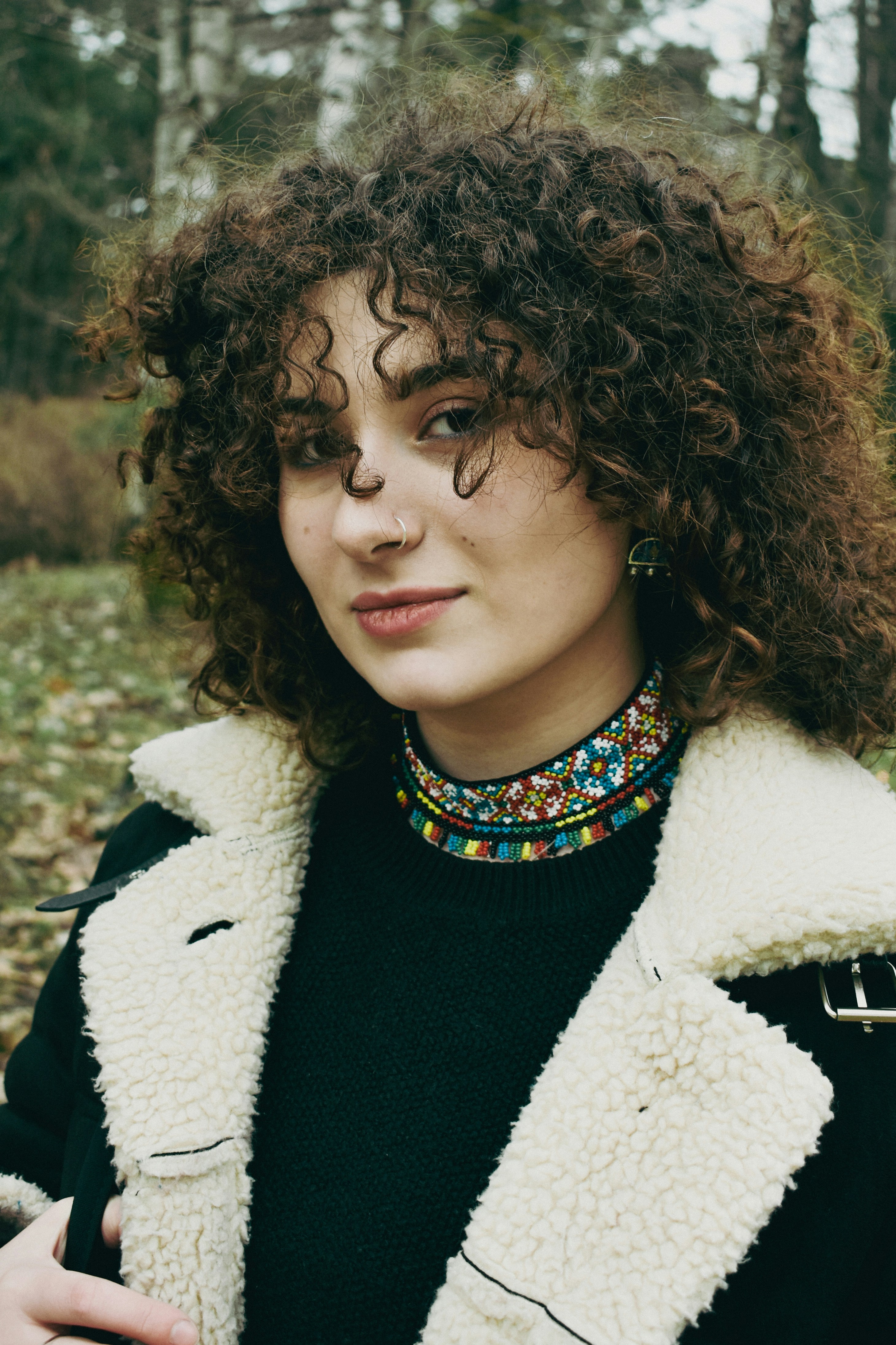 Young woman with curly hair wearing a colorful beaded necklace and a shearling coat, set against a natural backdrop of autumn foliage.