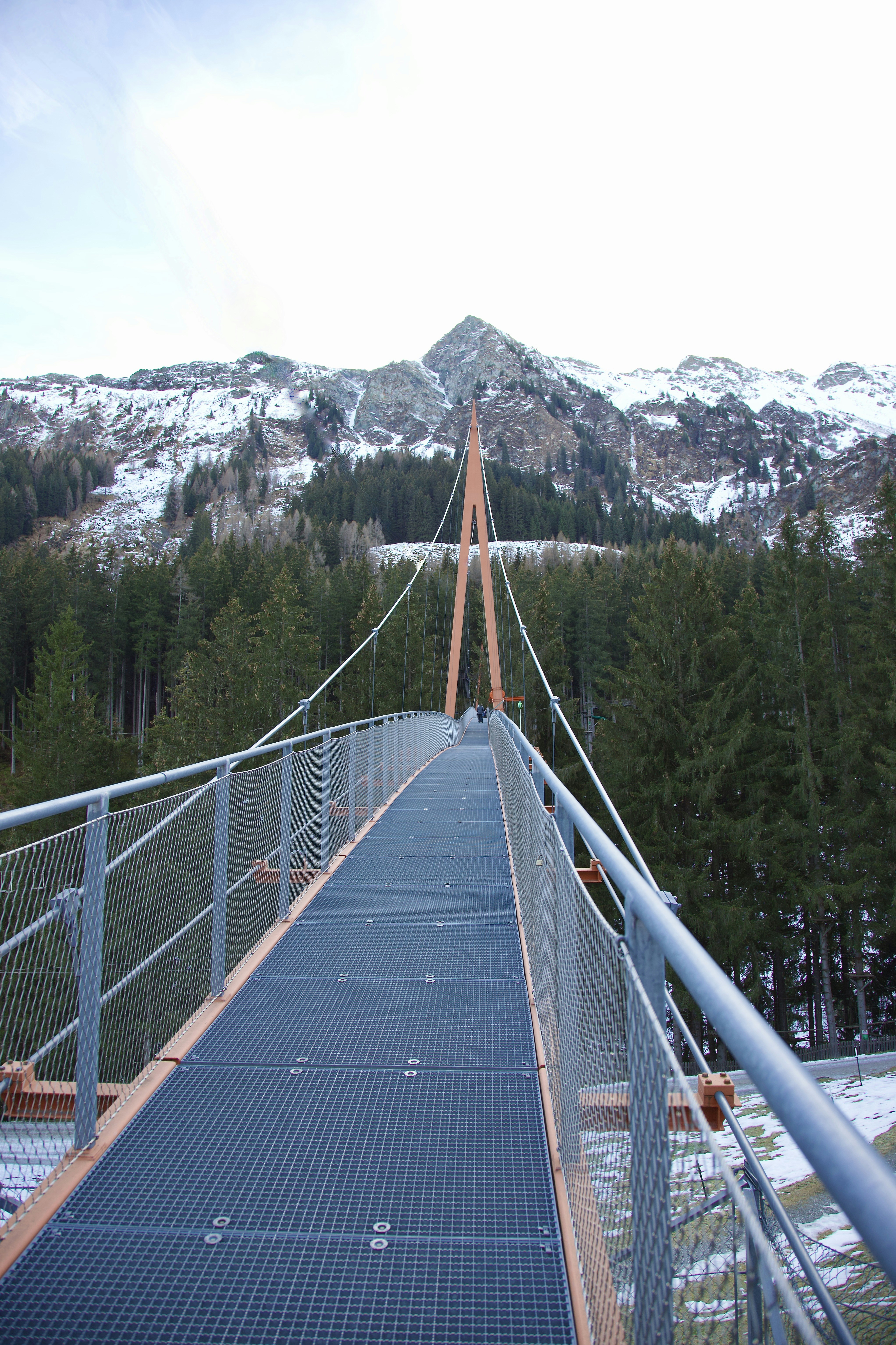 A suspension bridge stretches toward snow-capped mountains, framed by dense evergreen forests. The scene evokes a sense of exploration and tranquility.