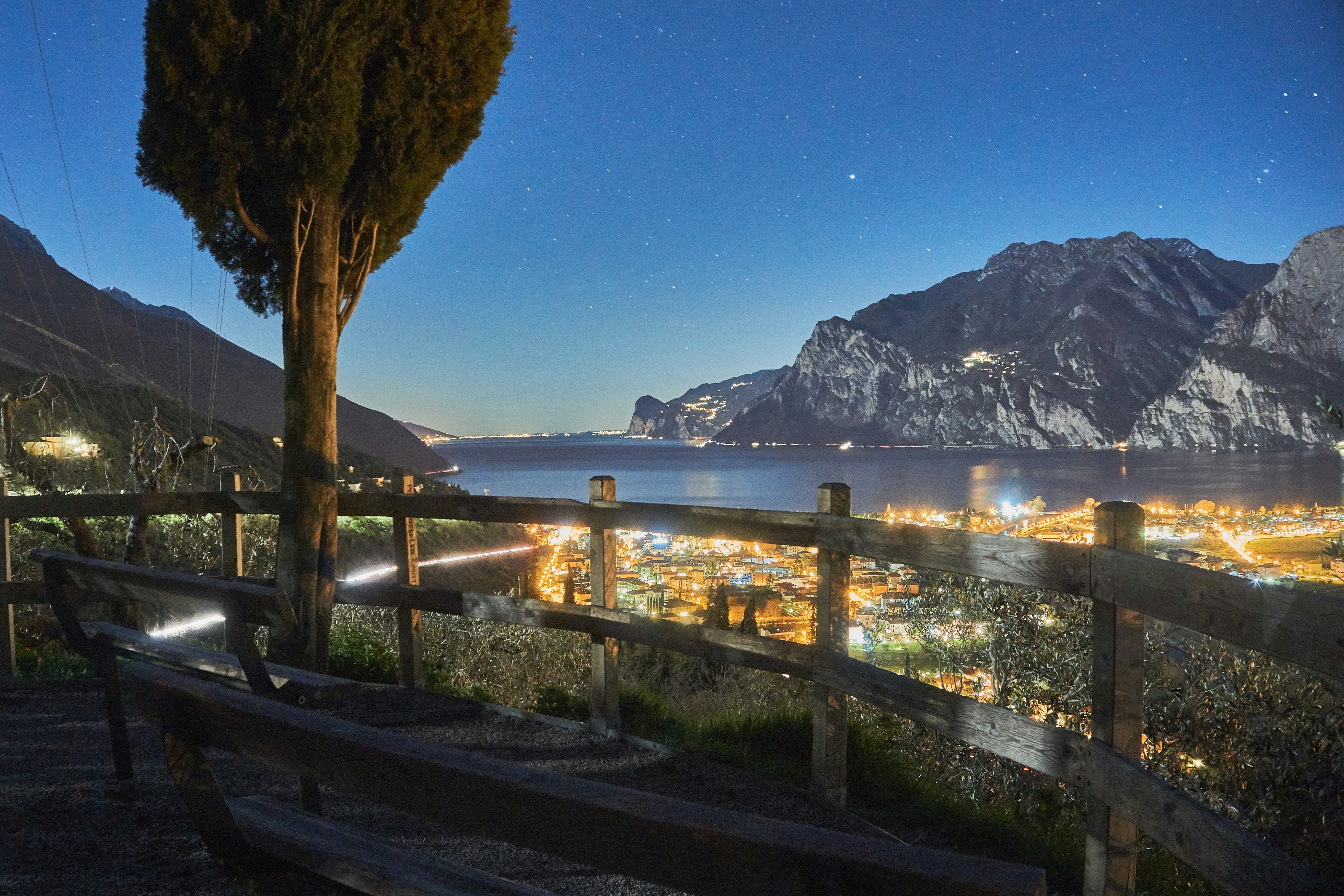 Wooden bench atop a hillside, overlooking a lit-up town by a mountain lake under a starry night sky.