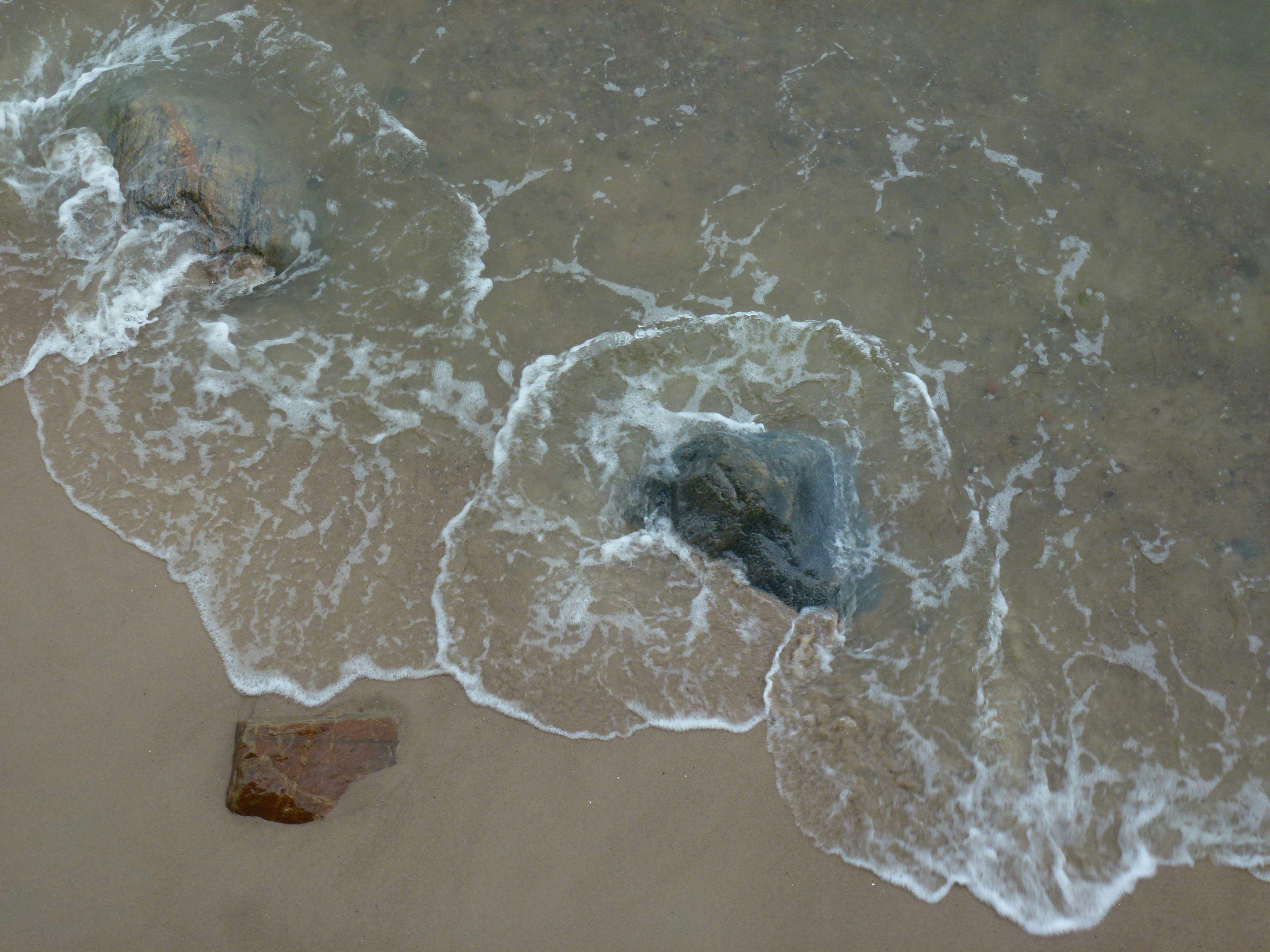 Dark rock sits within curling sea foam along a wet sandy shore, as waves wash around it. A small rusted fragment lies nearby on the sand.