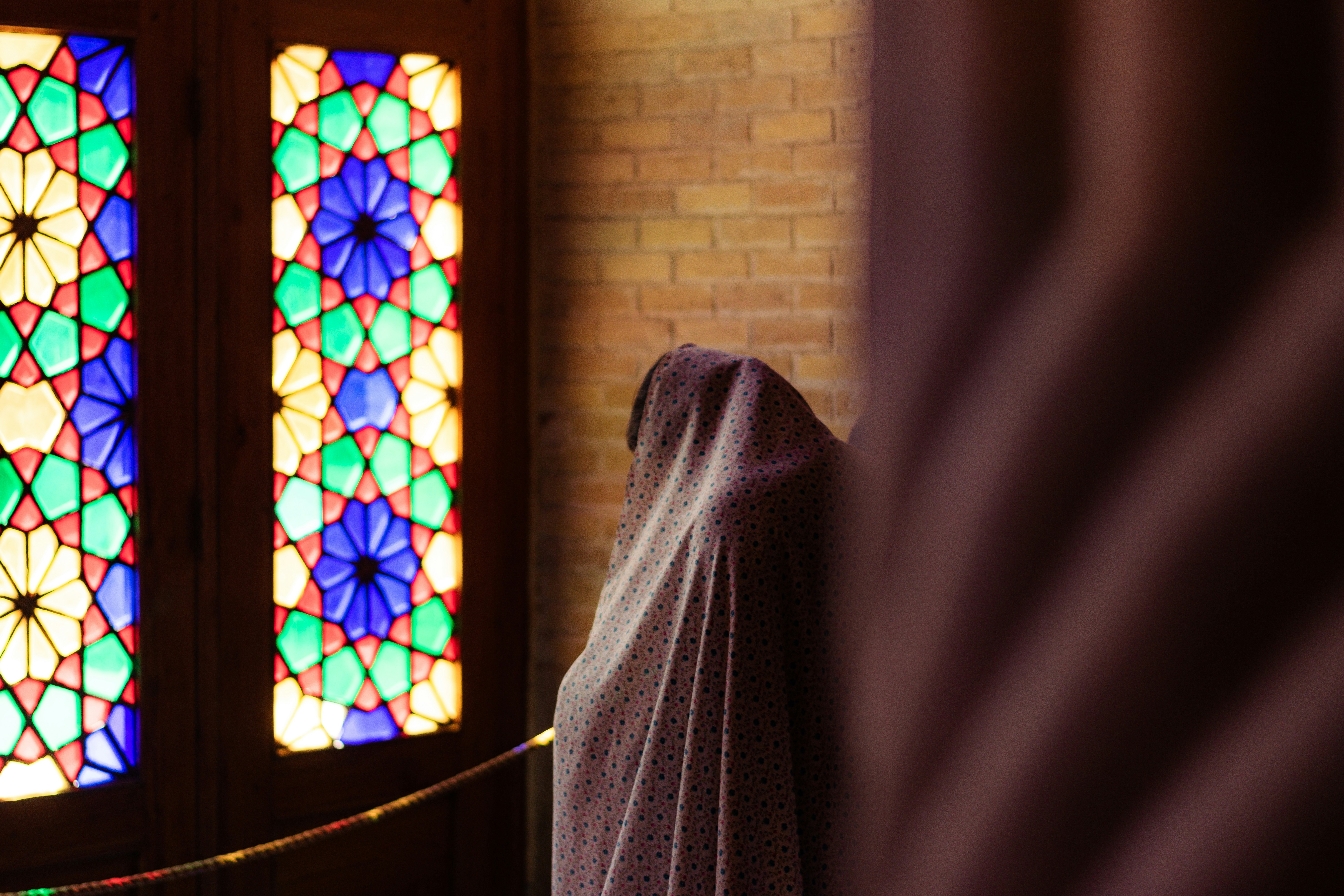 Figure draped in fabric standing beside vibrant stained glass windows in a dimly lit room.