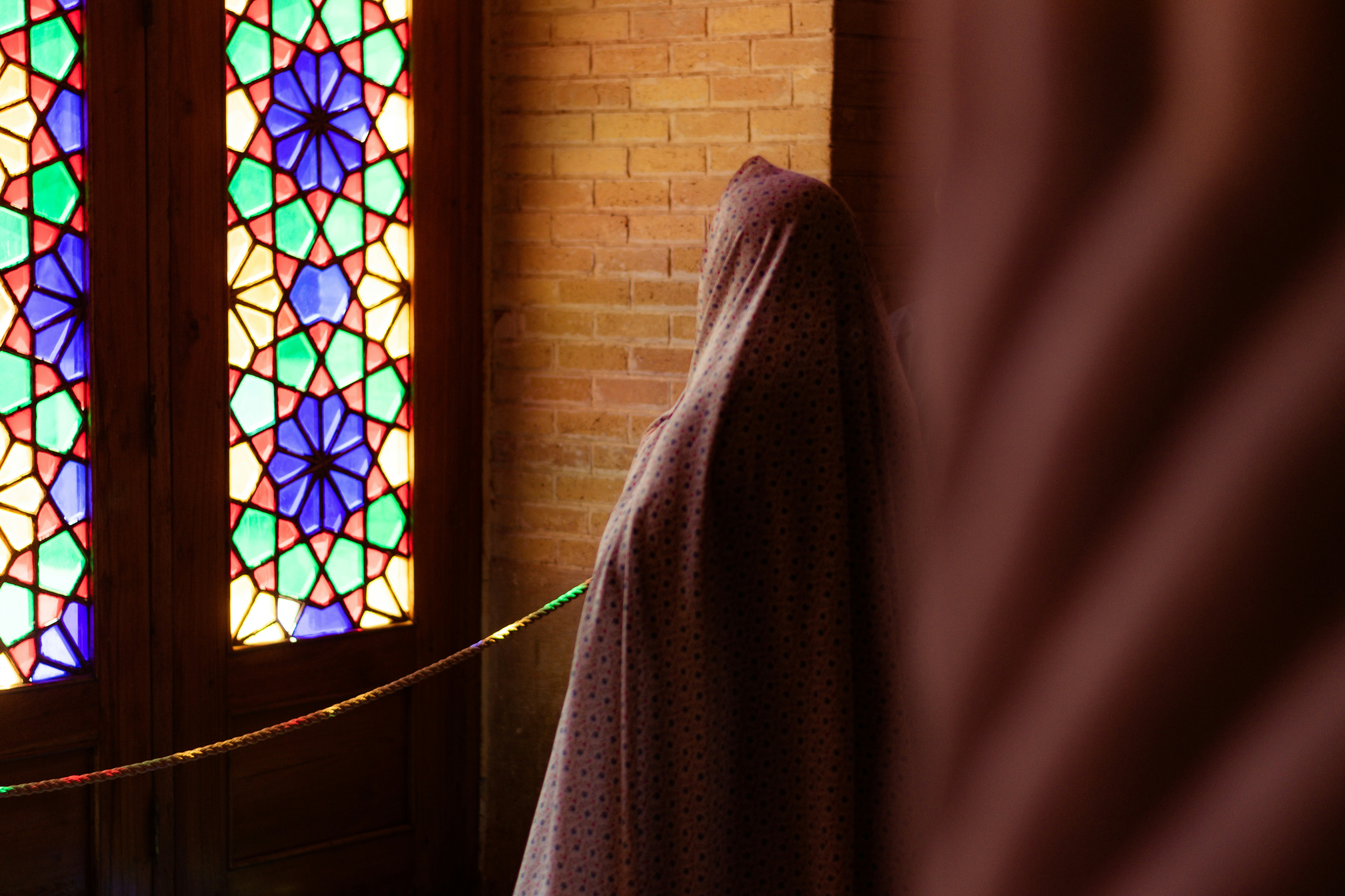 Figure shrouded in a cloak stands near colorful stained glass windows in a dimly lit space.