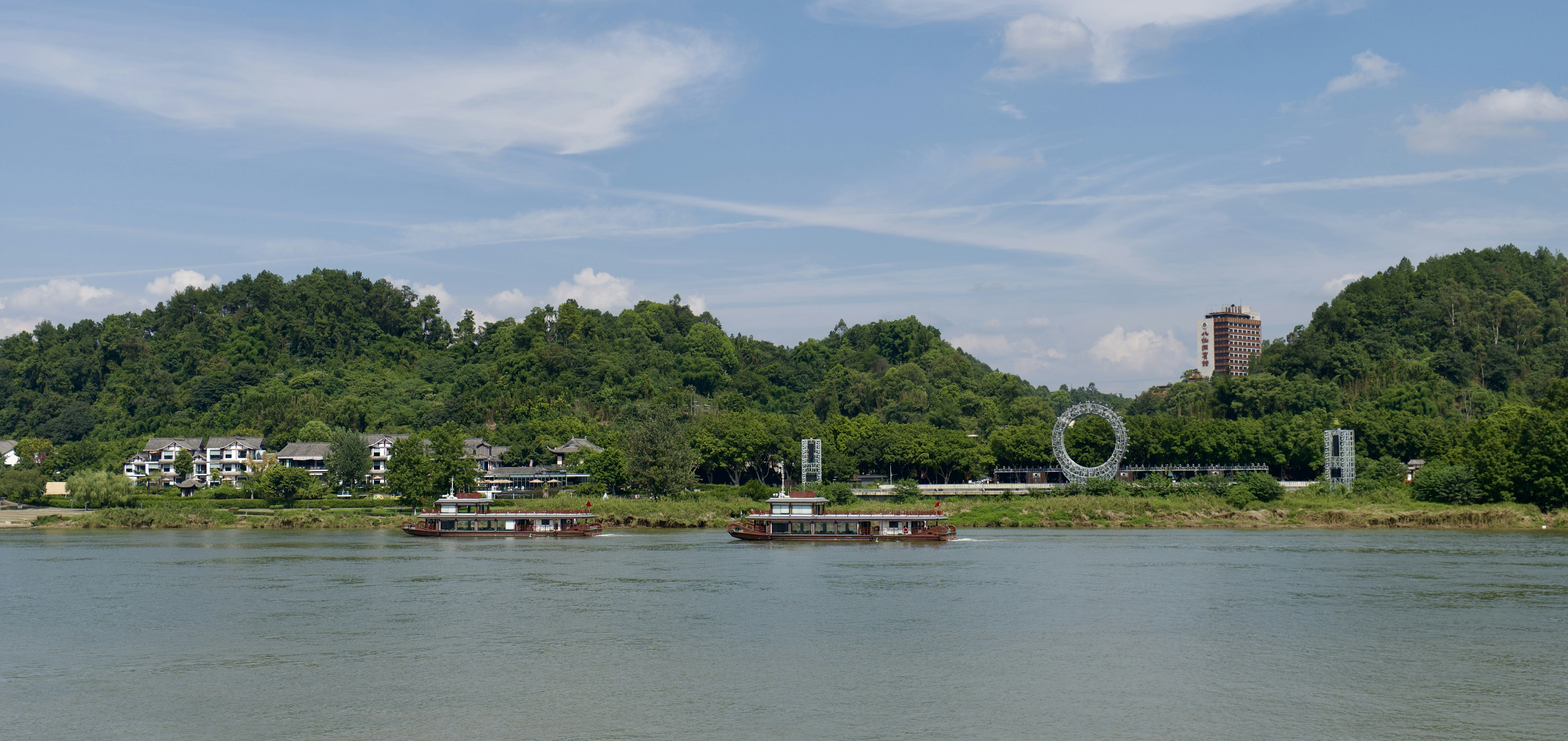 Serene riverfront with lush green hills and contemporary circular sculptures under a bright blue sky.