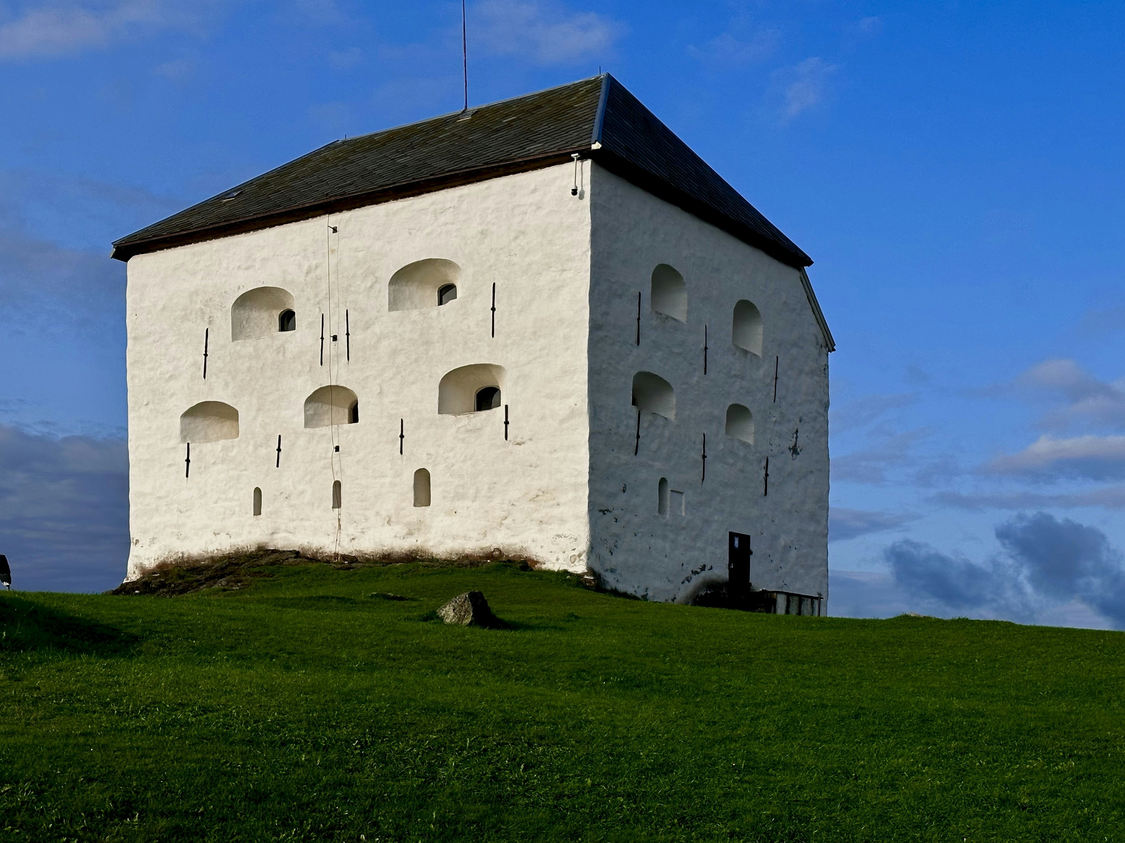 White stone building with arched windows on a grassy hill under a bright blue sky.