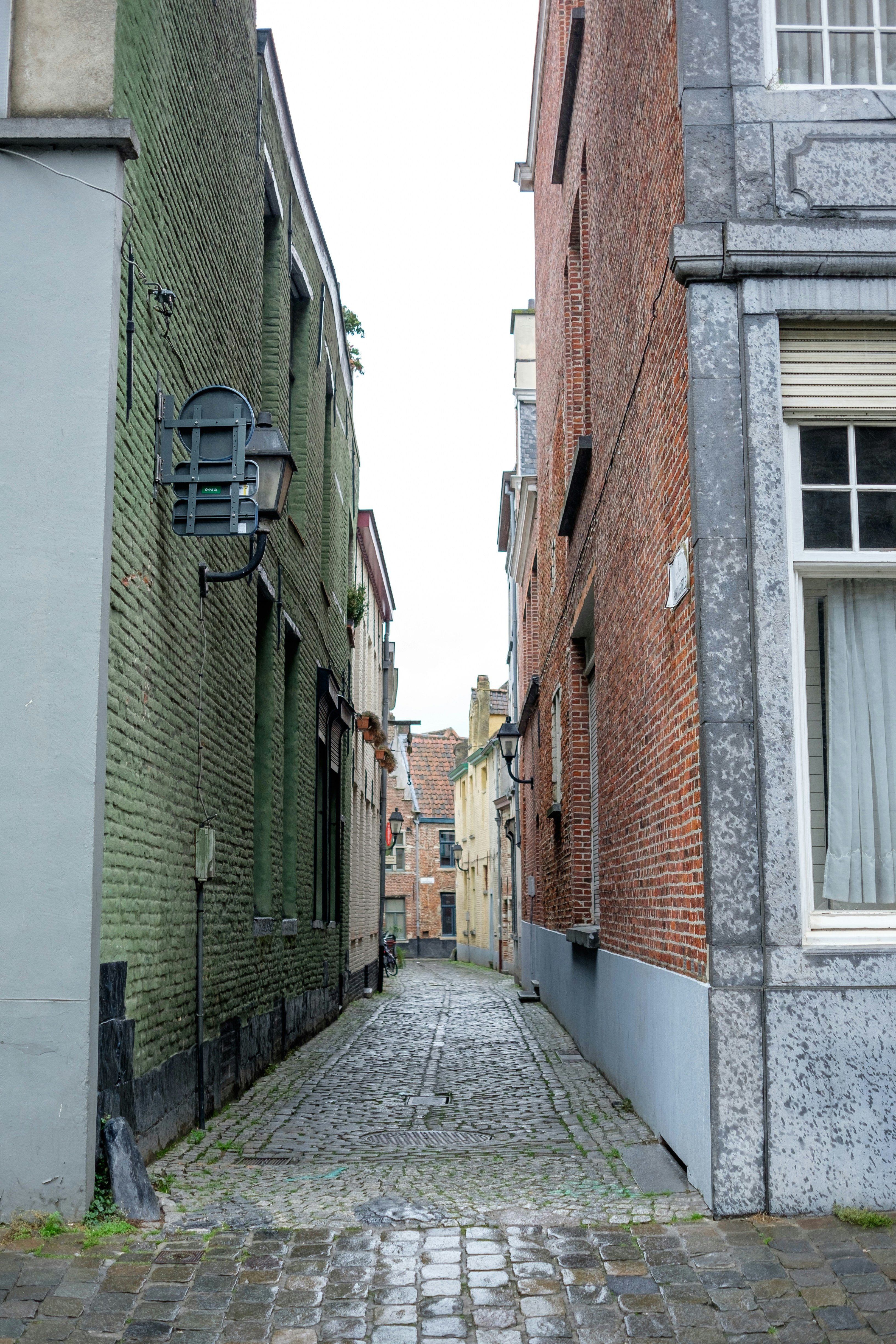 Narrow cobblestone alley flanked by textured green and red brick walls, leading to a distant view of quaint buildings. Wet surfaces reflect the muted light.