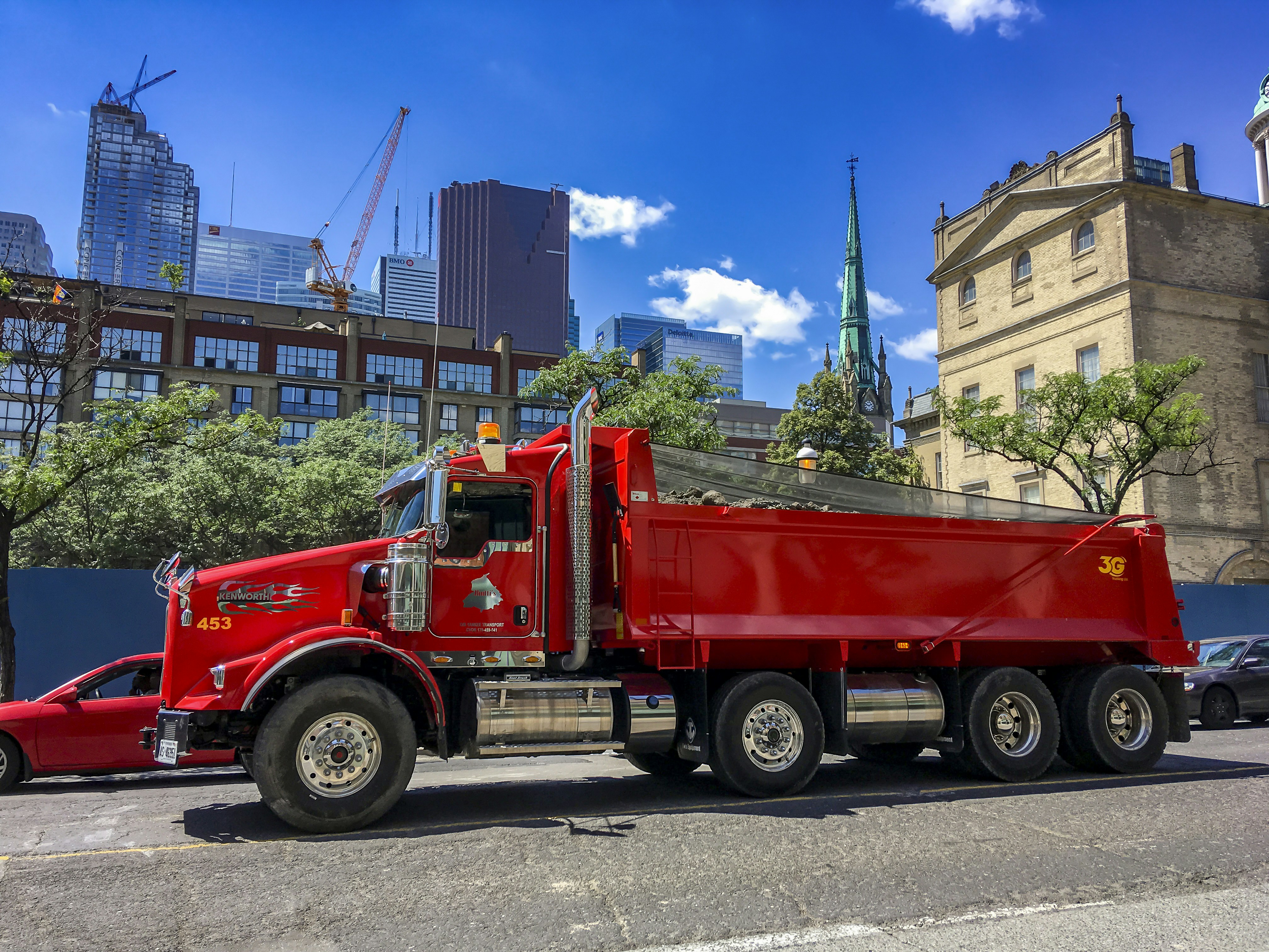 Red dump truck driving alongside tall city buildings under a clear blue sky.