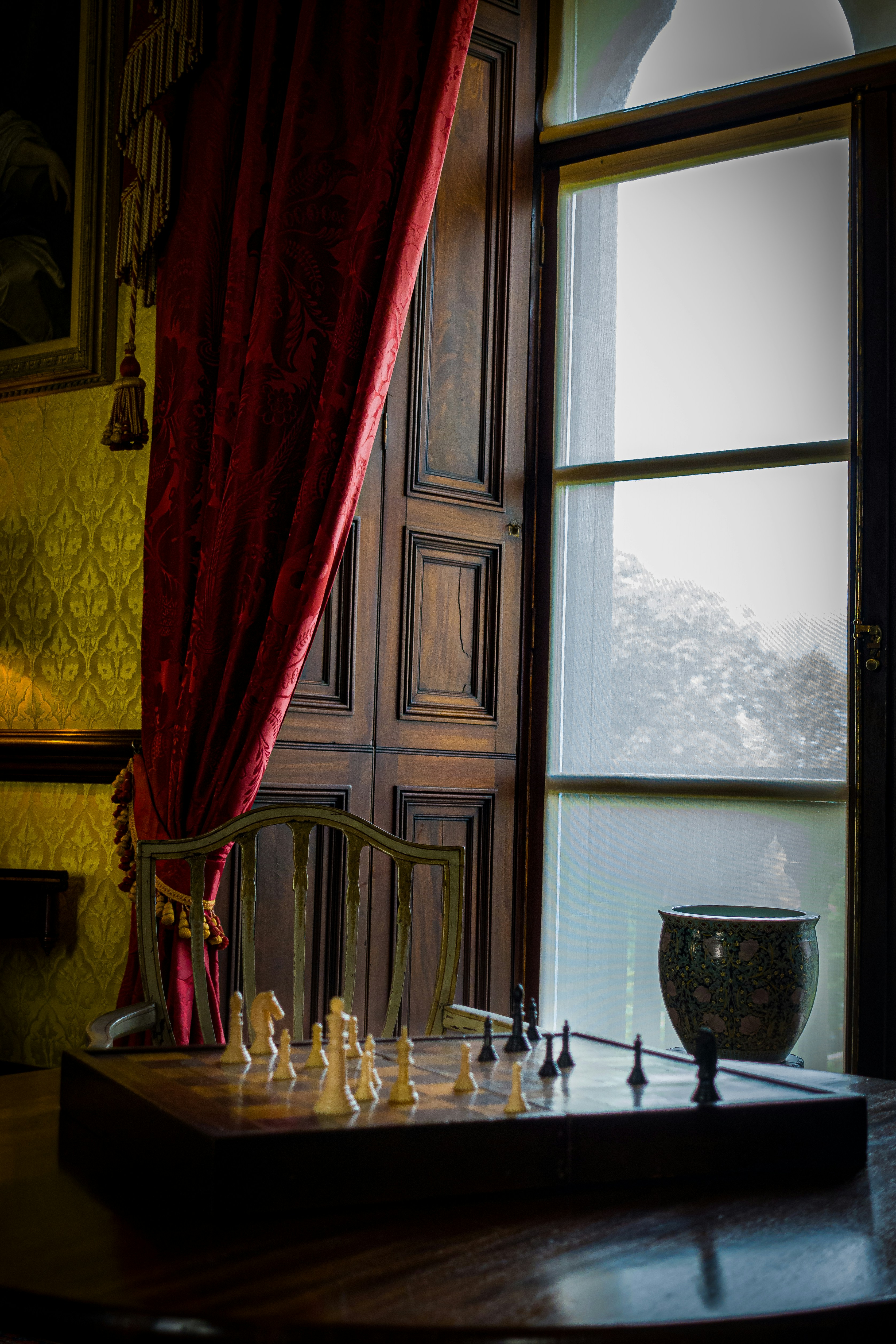 Photograph of a chessboard with scattered pieces on a dark wooden table beside a red velvet curtain and a bright window. The composition emphasizes still-life contrasts within a vintage parlor.
