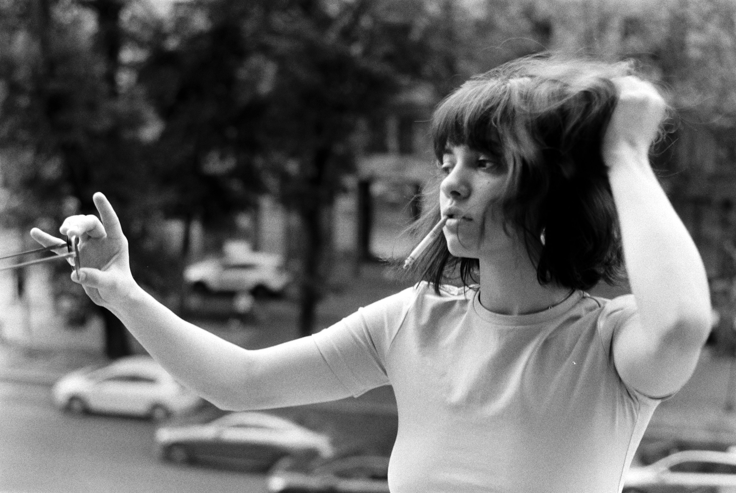 The black-and-white image captures a young woman in a candid moment as she adjusts her hair, with a cigarette poised between her lips. The composition draws focus to her serene expression against a blurred urban backdrop, evoking a sense of introspection. The high contrast and soft lighting enhance the mood, creating a visually striking interplay between the subject and her surroundings.