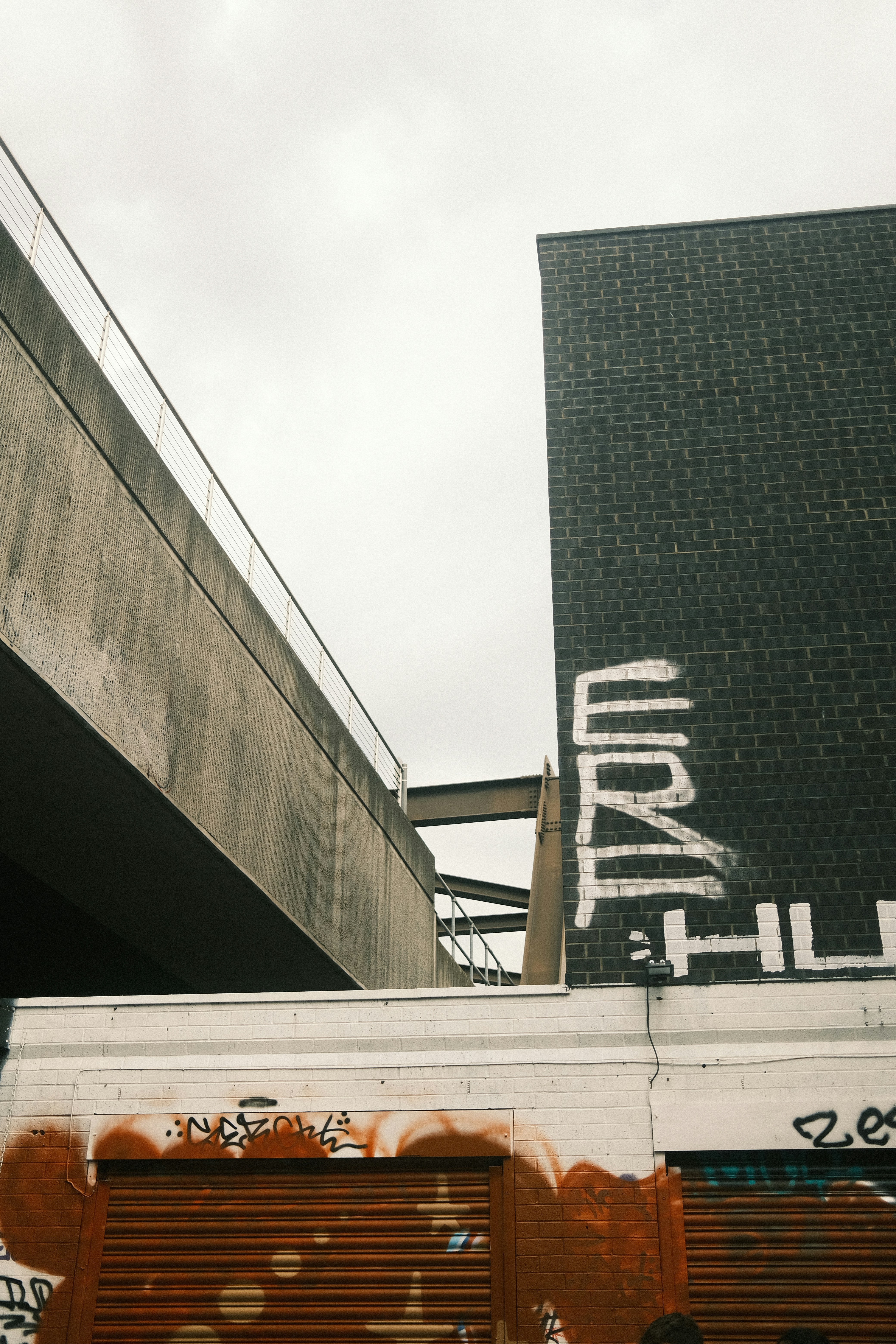 Vertical Glyphs on Brick: White Letters Ascend the City Facade Under an Industrial Overpass