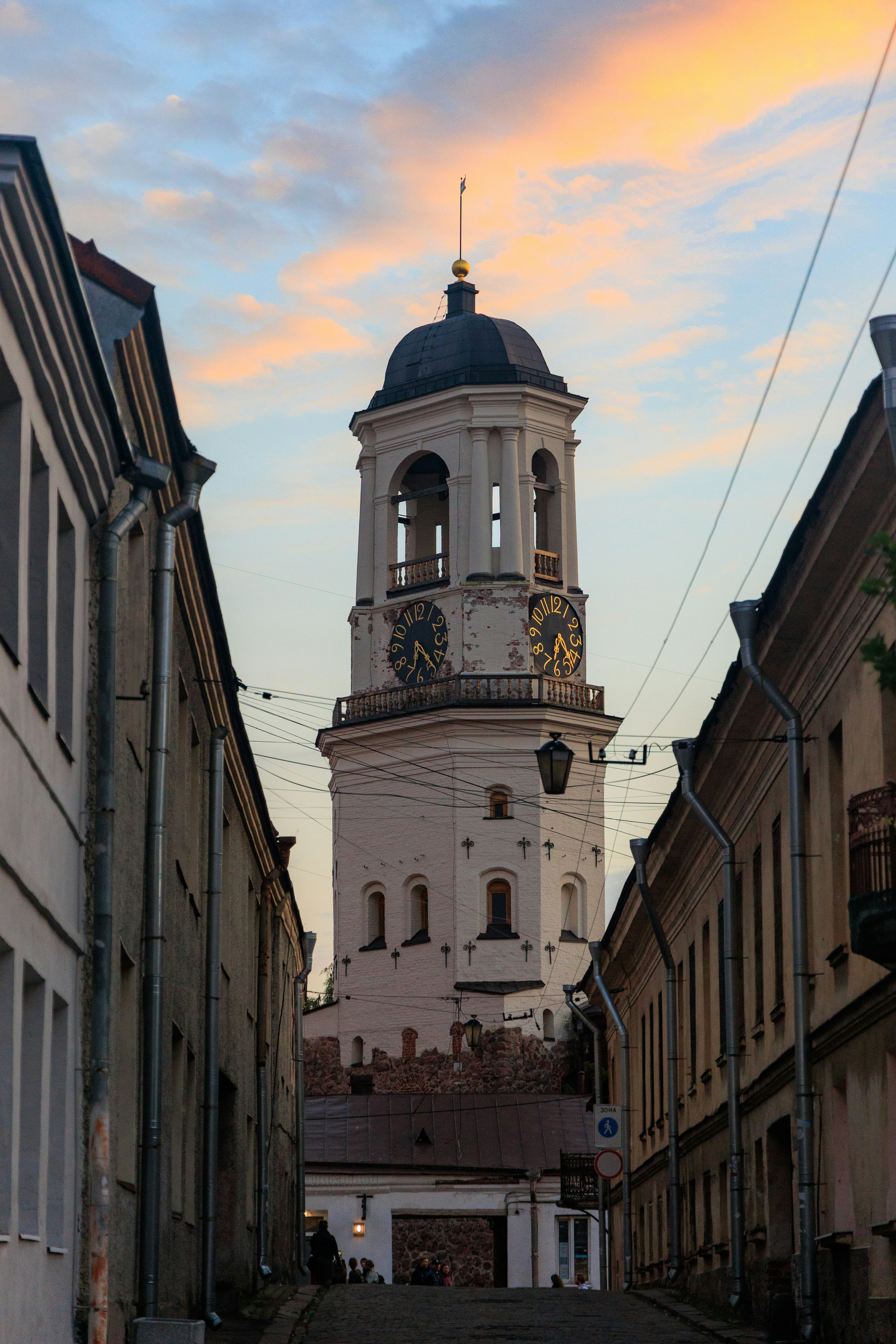 Historic clock tower rises above narrow cobblestone street at twilight, framed by charming old buildings. 