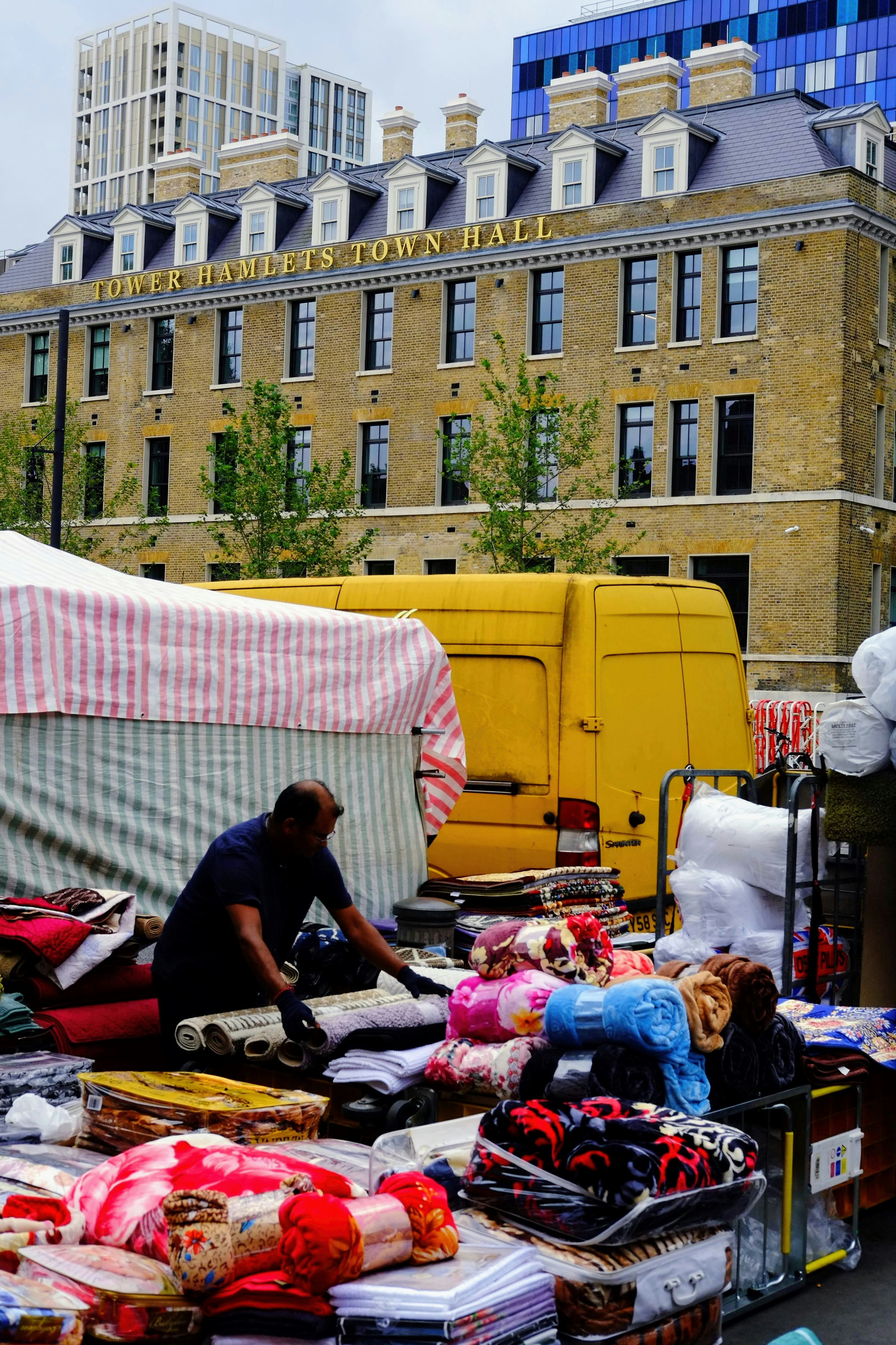 A bustling street market scene with vendors sorting colorful blankets and rugs beside a bright yellow van, set against the brick façade of Tower Hamlets Town Hall.