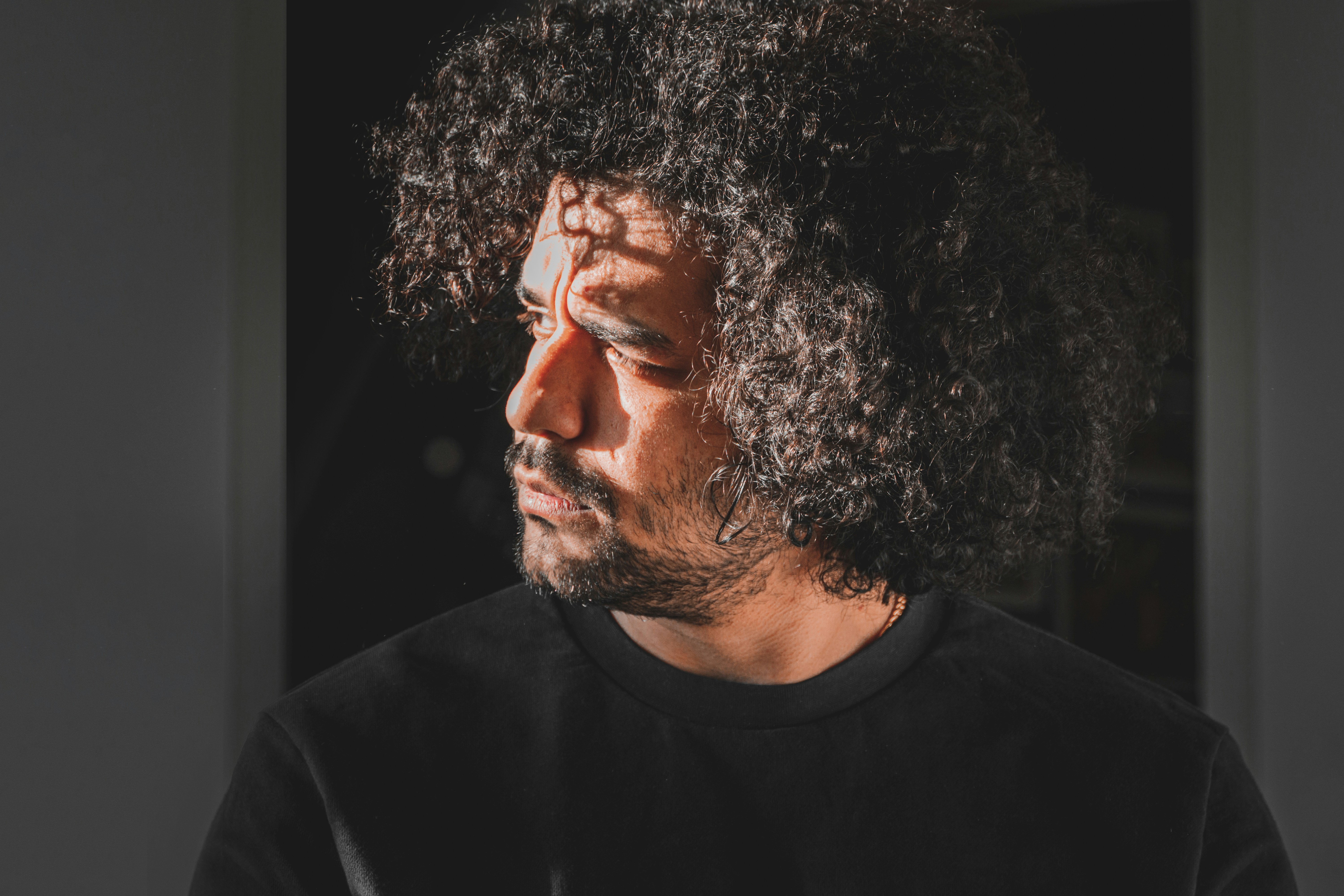 Man with curly hair gazing thoughtfully, illuminated by soft light that highlights his features against a dark background.