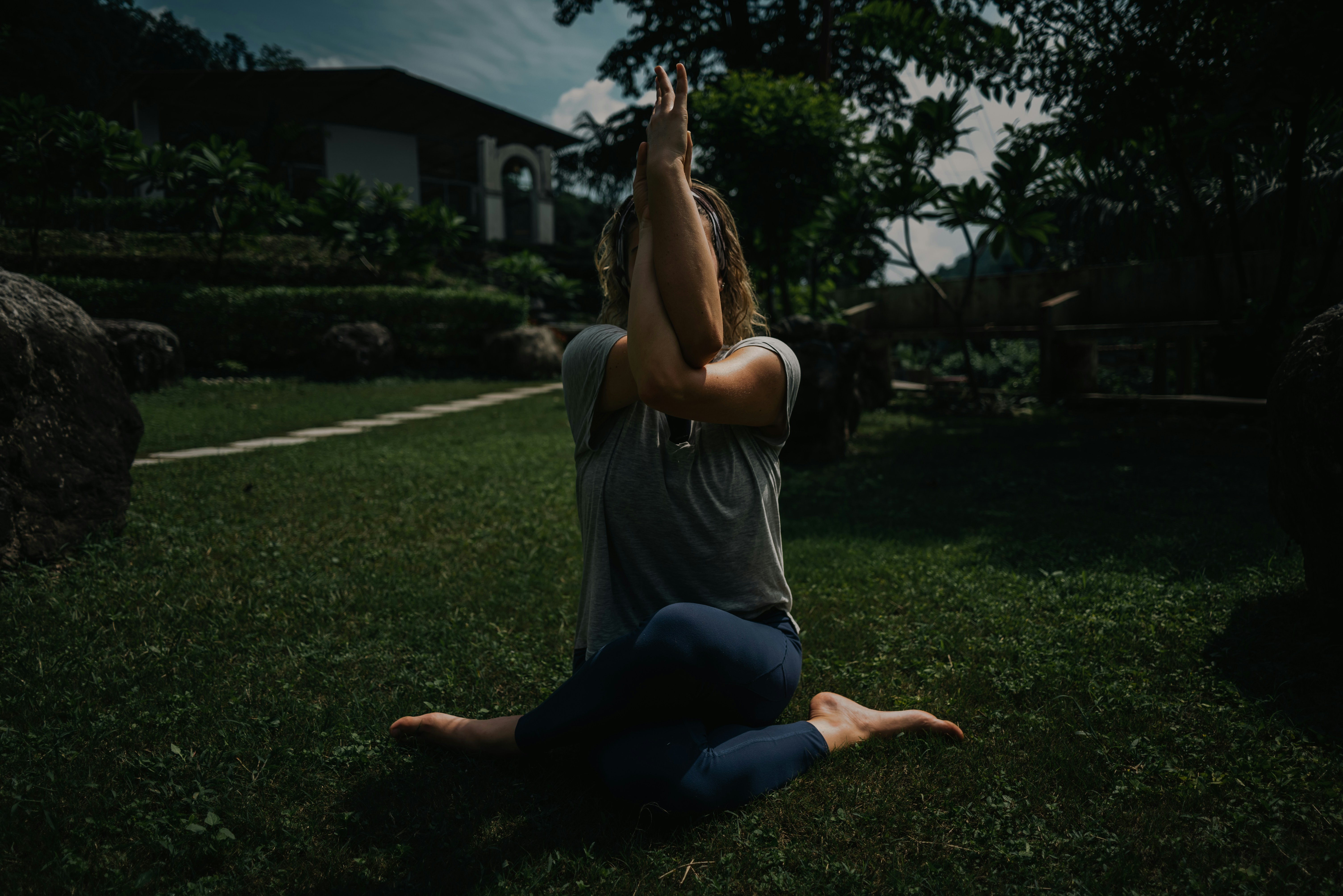 Person practicing yoga in a lush garden with dappled sunlight and a house in the background.