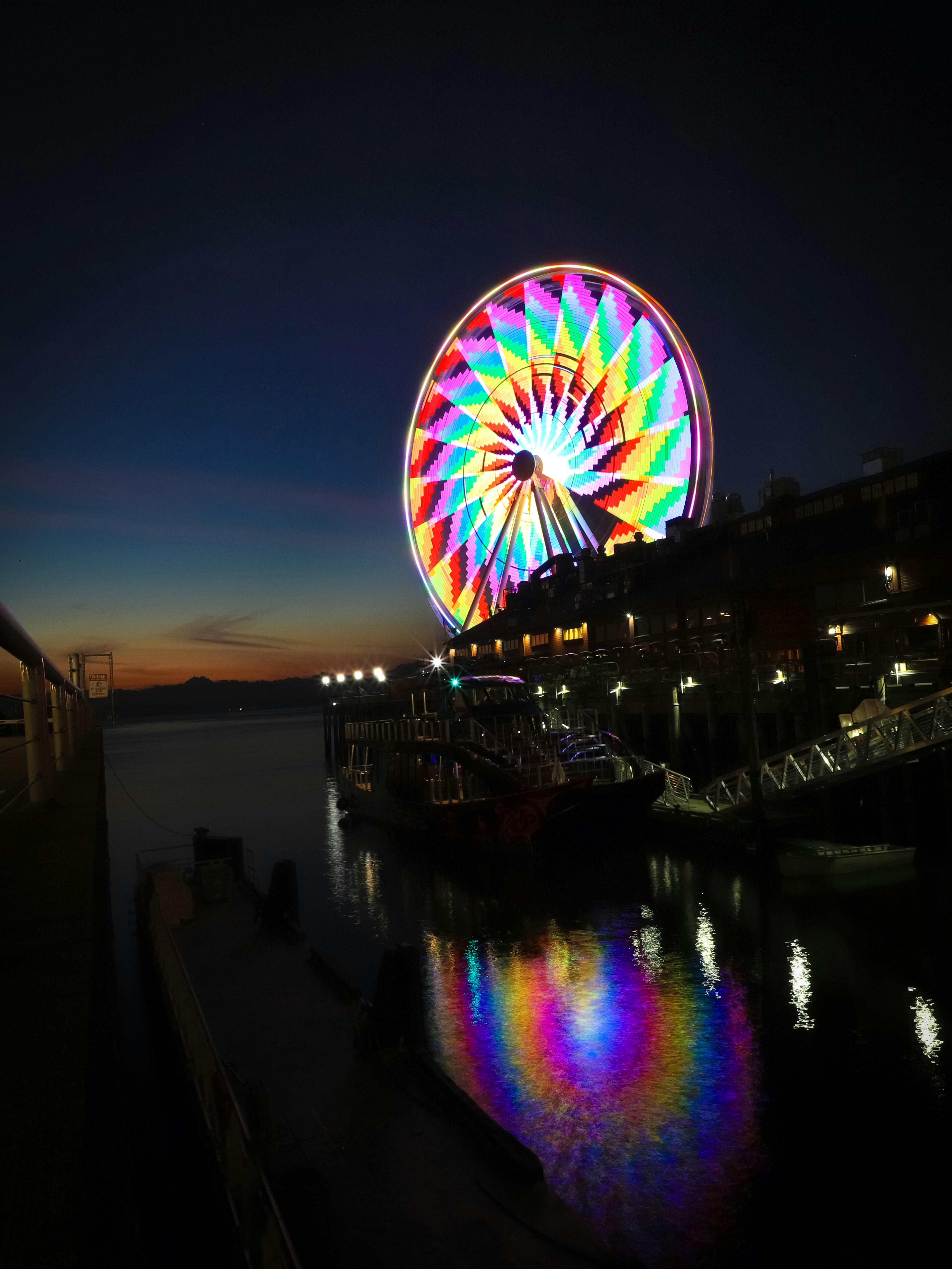 Colorful ferris wheel lights reflecting on the water at dusk, with a silhouetted pier in the foreground.