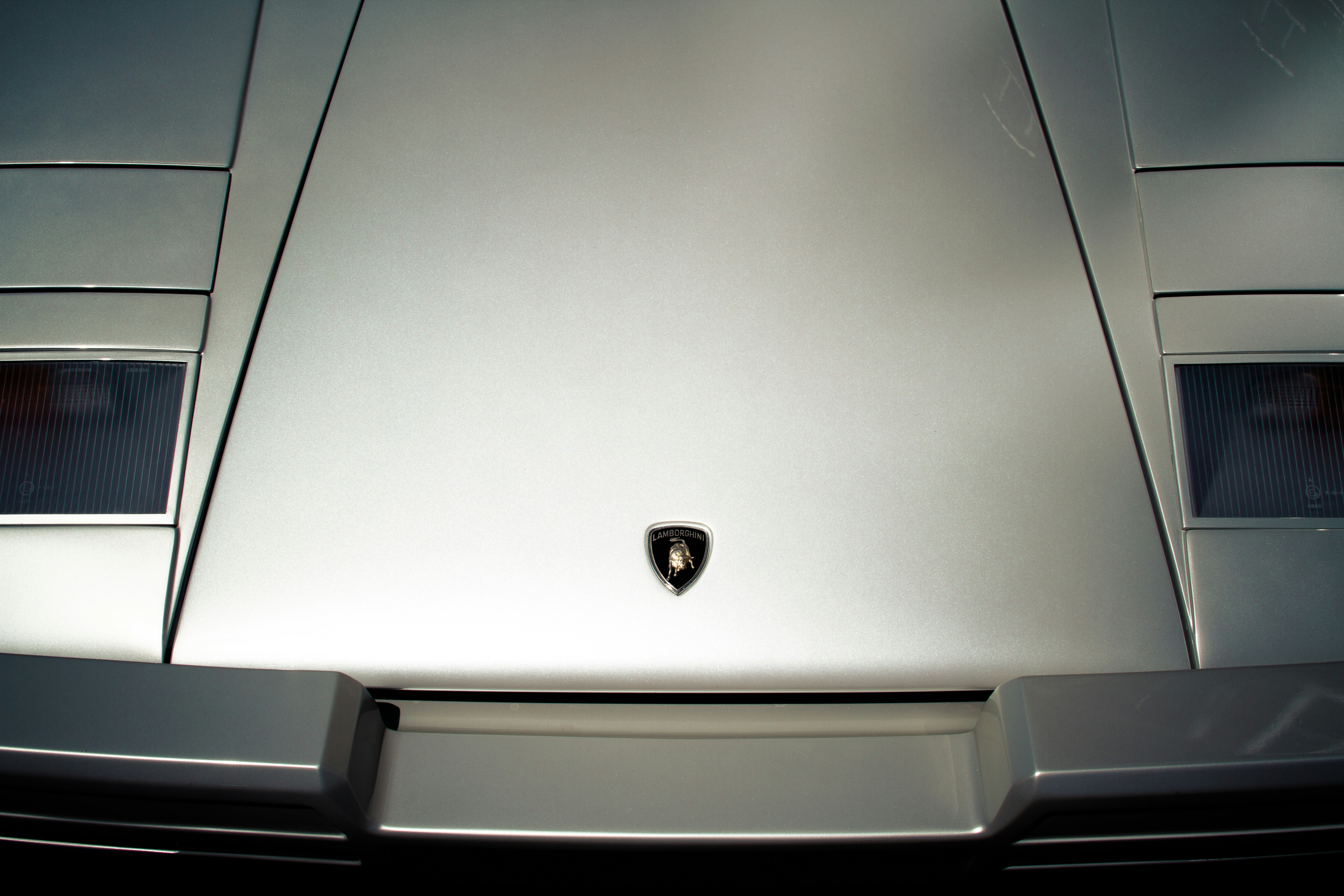 Sleek, silver Lamborghini hood showcasing the iconic emblem and angular design. The polished surface reflects light, emphasizing its aerodynamic shape.