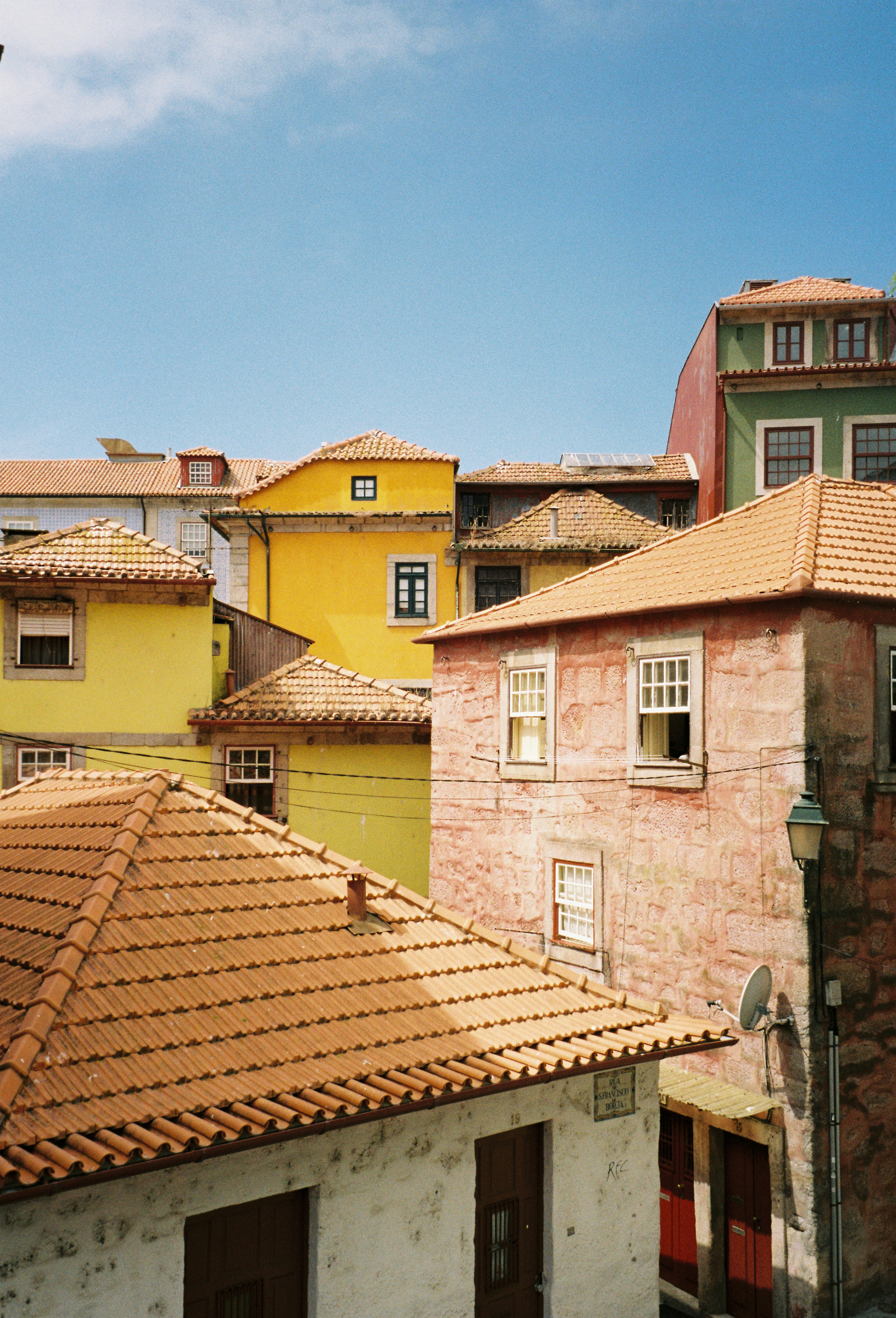 Colorful Mediterranean-style houses with terracotta roofs stack along a sunlit street under a bright blue sky.