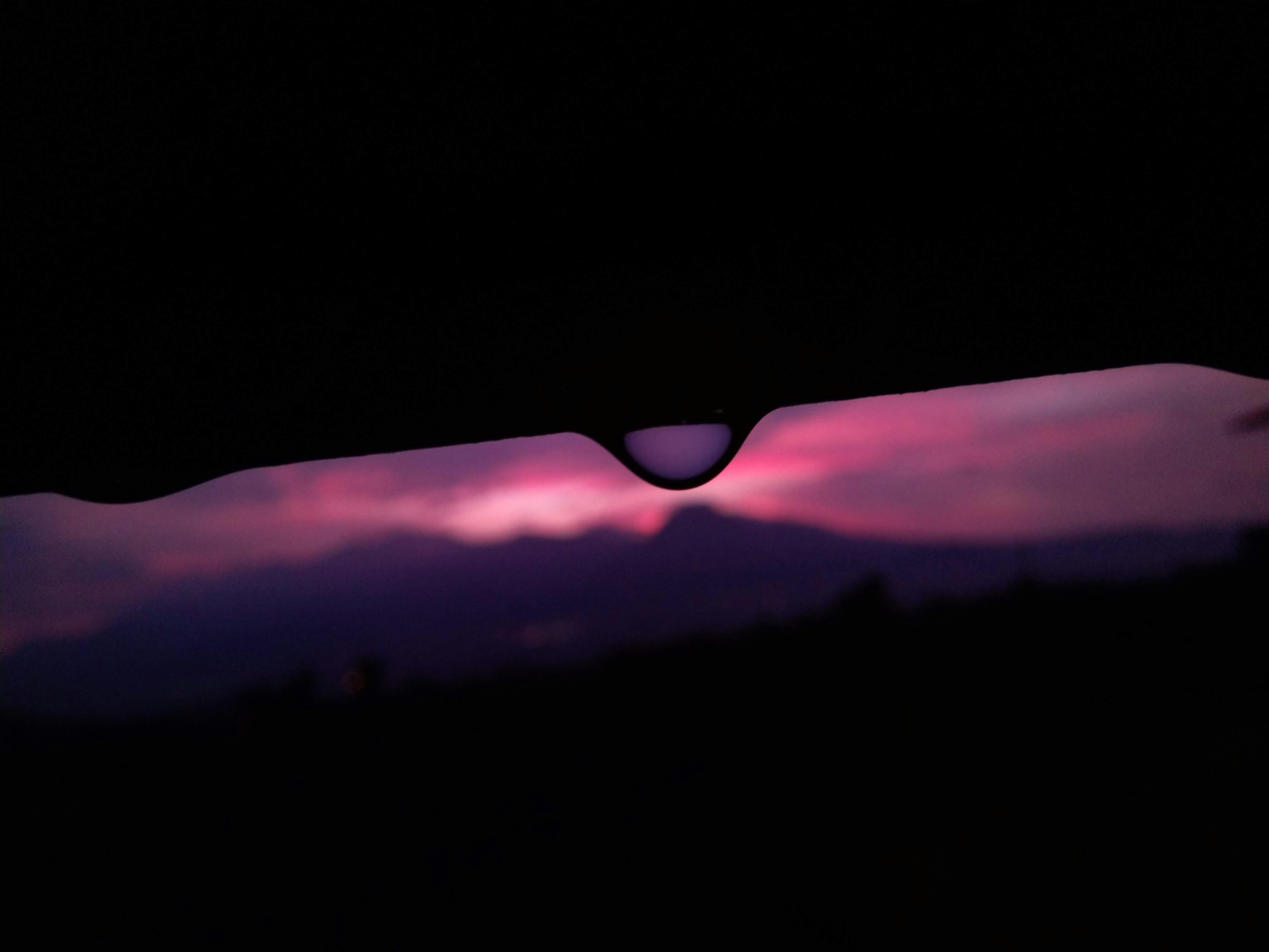 A photograph of a dew droplet perched on a curved edge against a dark silhouette and a pink-hued sunset. The composition emphasizes minimalism and mood.