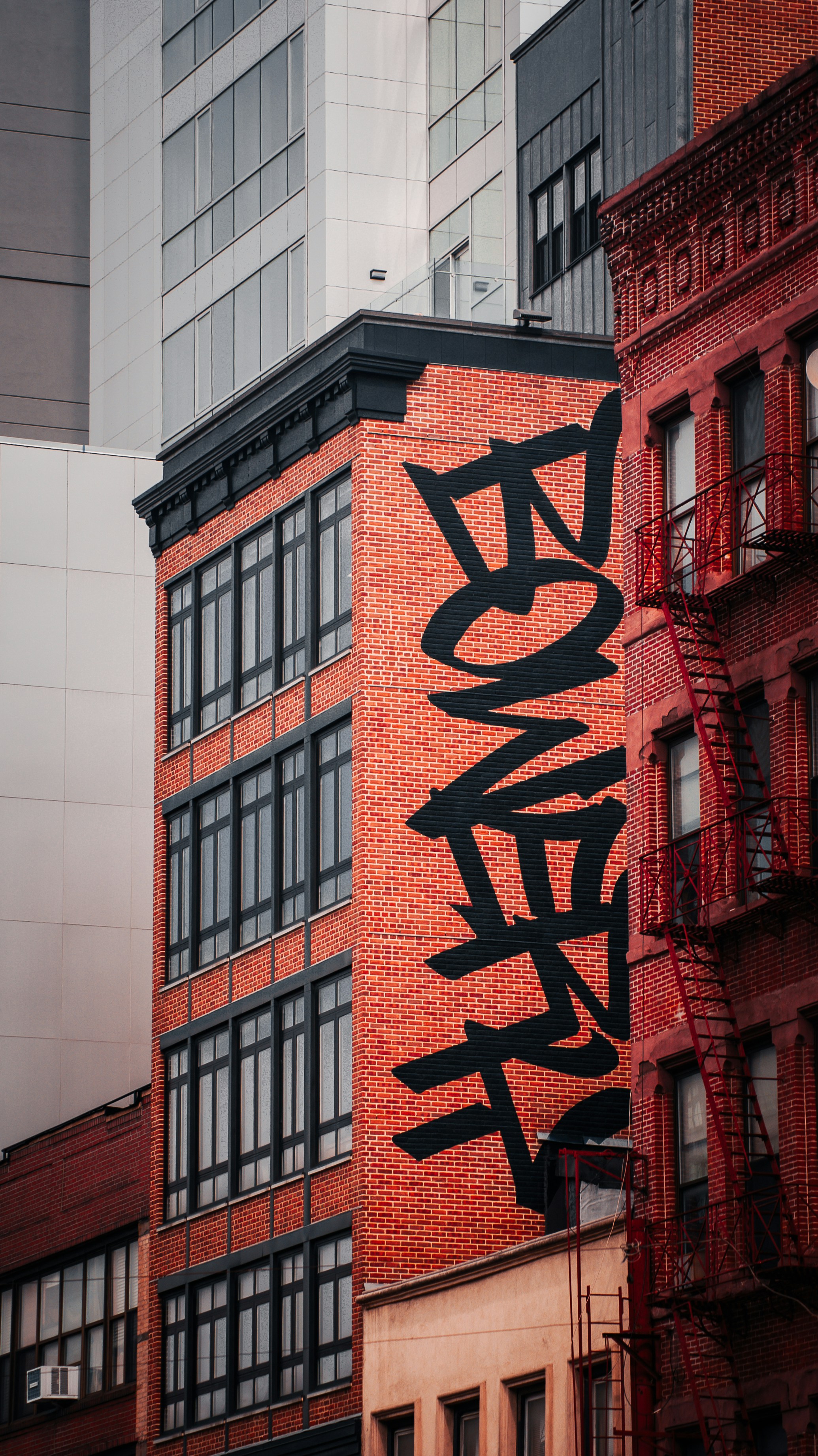 Tall red brick building features oversized vertical black graffiti characters across its facade beside a modern glass-and-concrete structure.