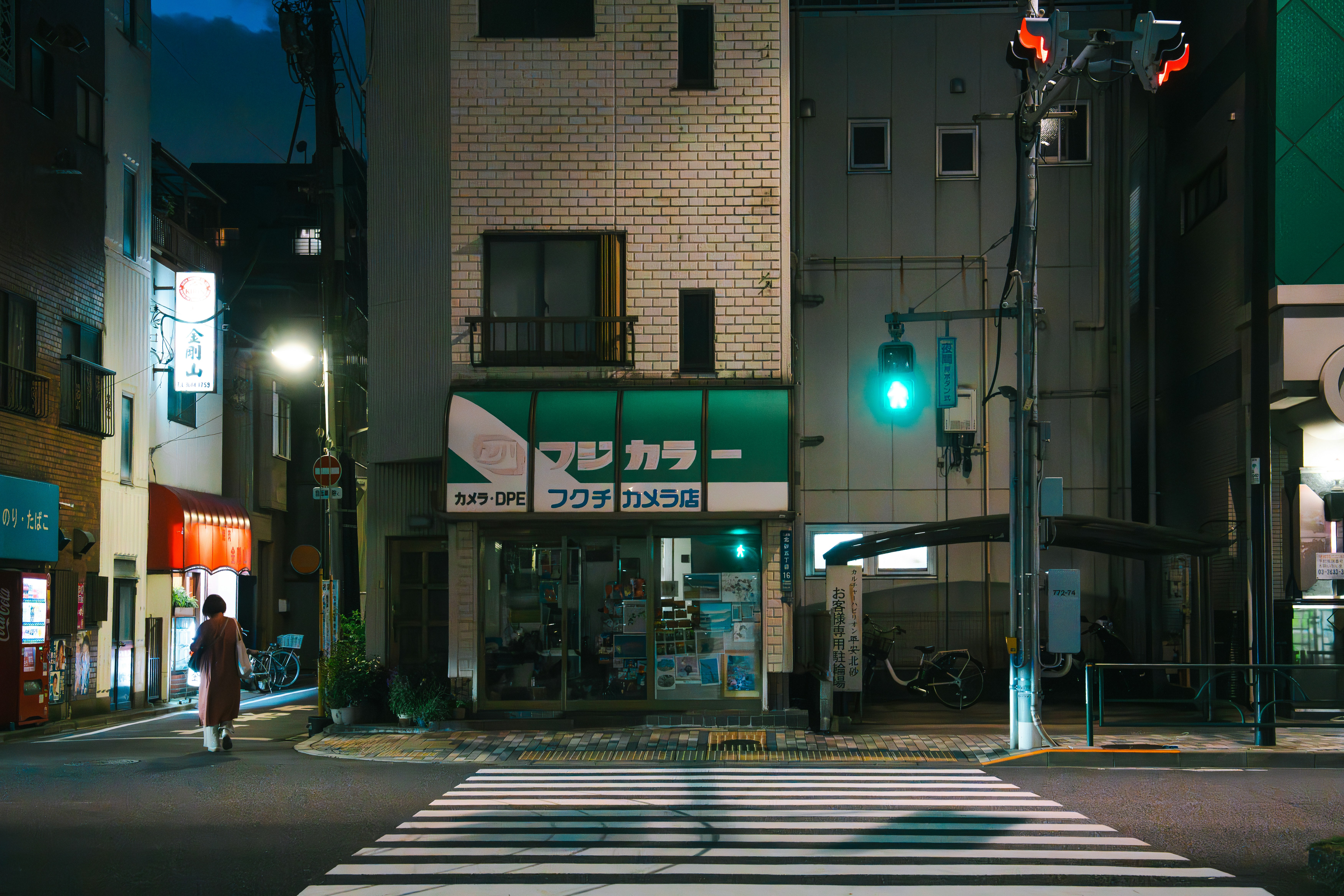A lone figure walks past a small storefront under a green traffic light in a quiet urban setting at night.