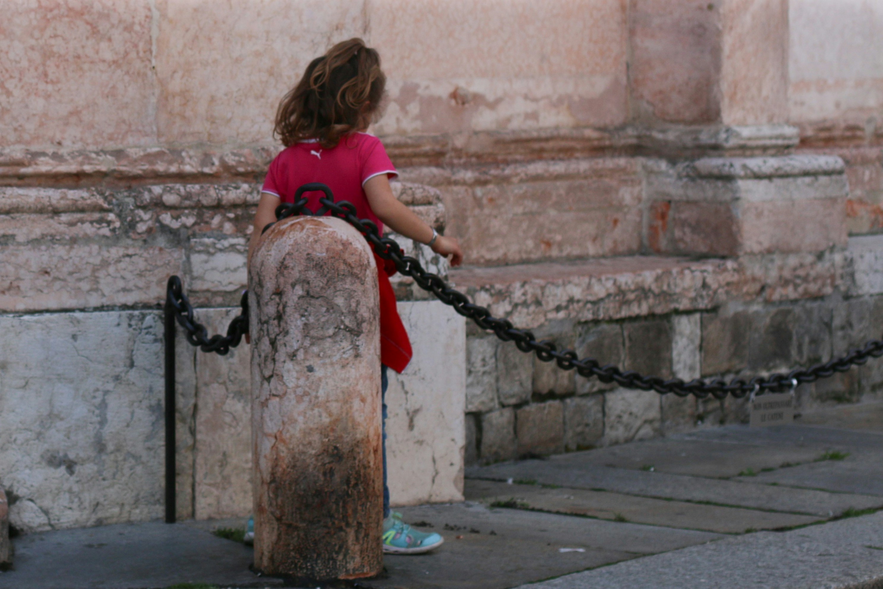 Young girl in a red shirt interacts with a chain barrier near a stone pillar on a city street.