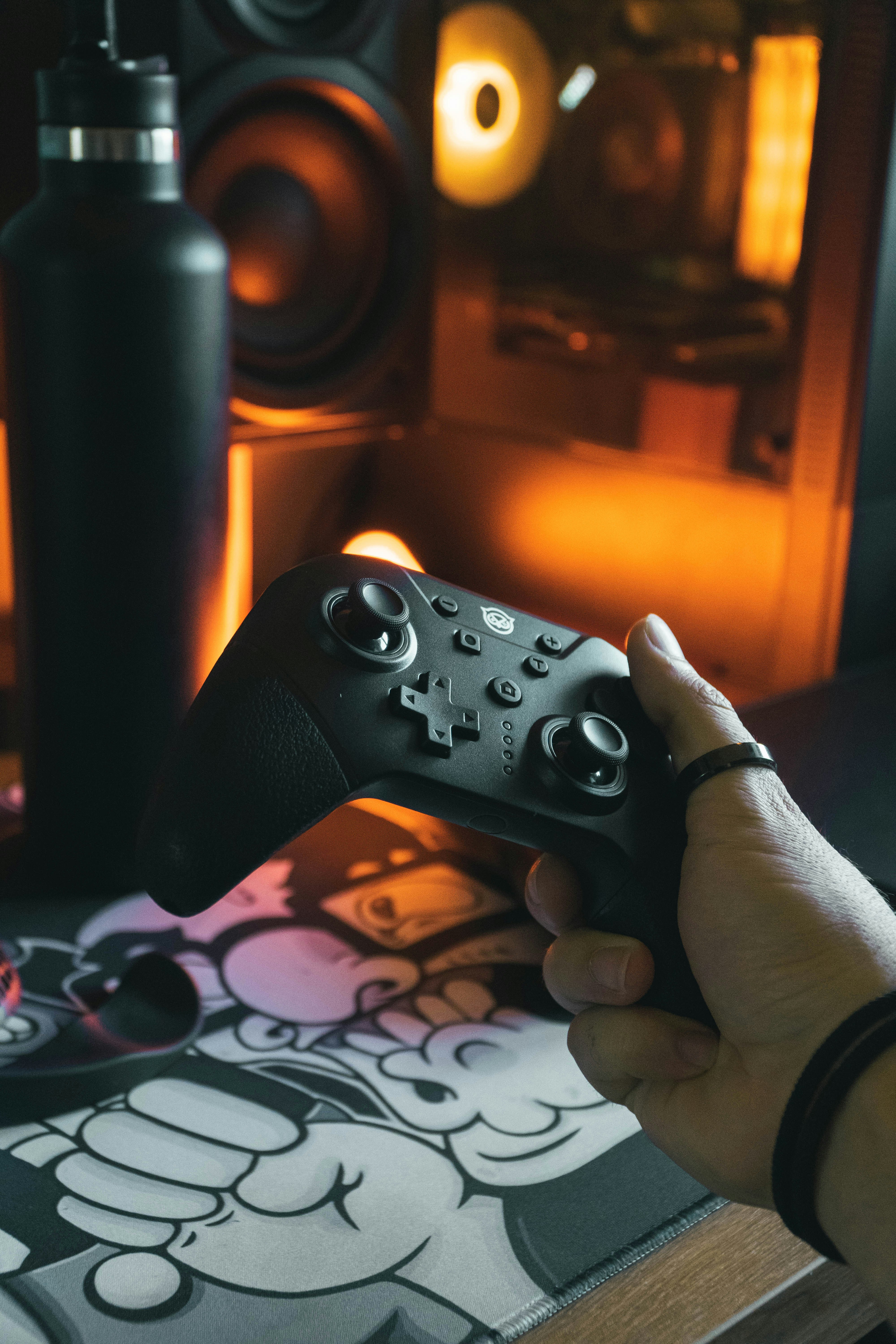 A hand gripping a sleek black game controller in a dimly lit gaming setup, with vibrant ambient lighting in the background.