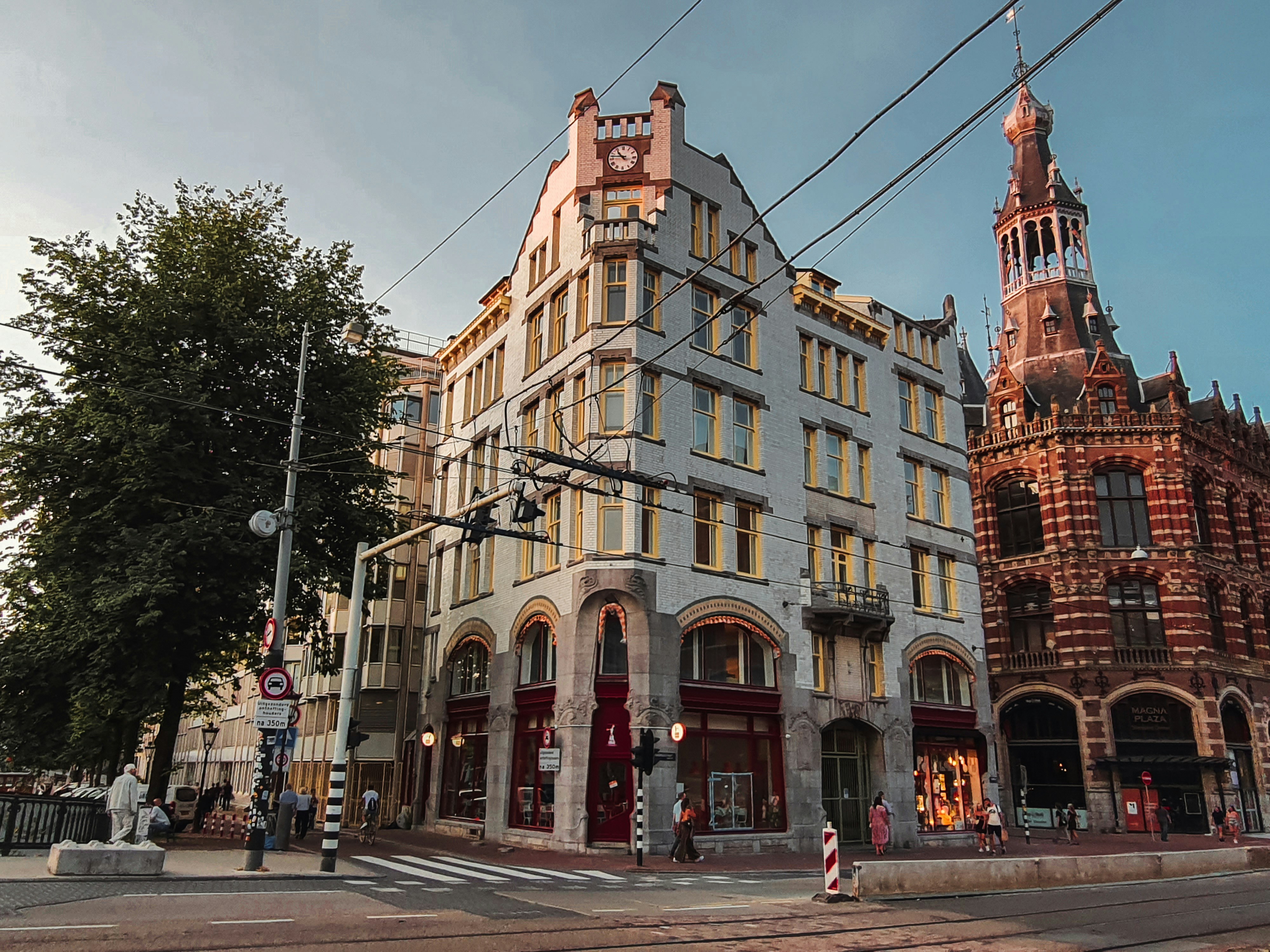 Pastel-facade buildings line a busy city street, with a distinctive red-brick tower rising on the right. A daytime urban photograph capturing pedestrians and storefronts along the corner.