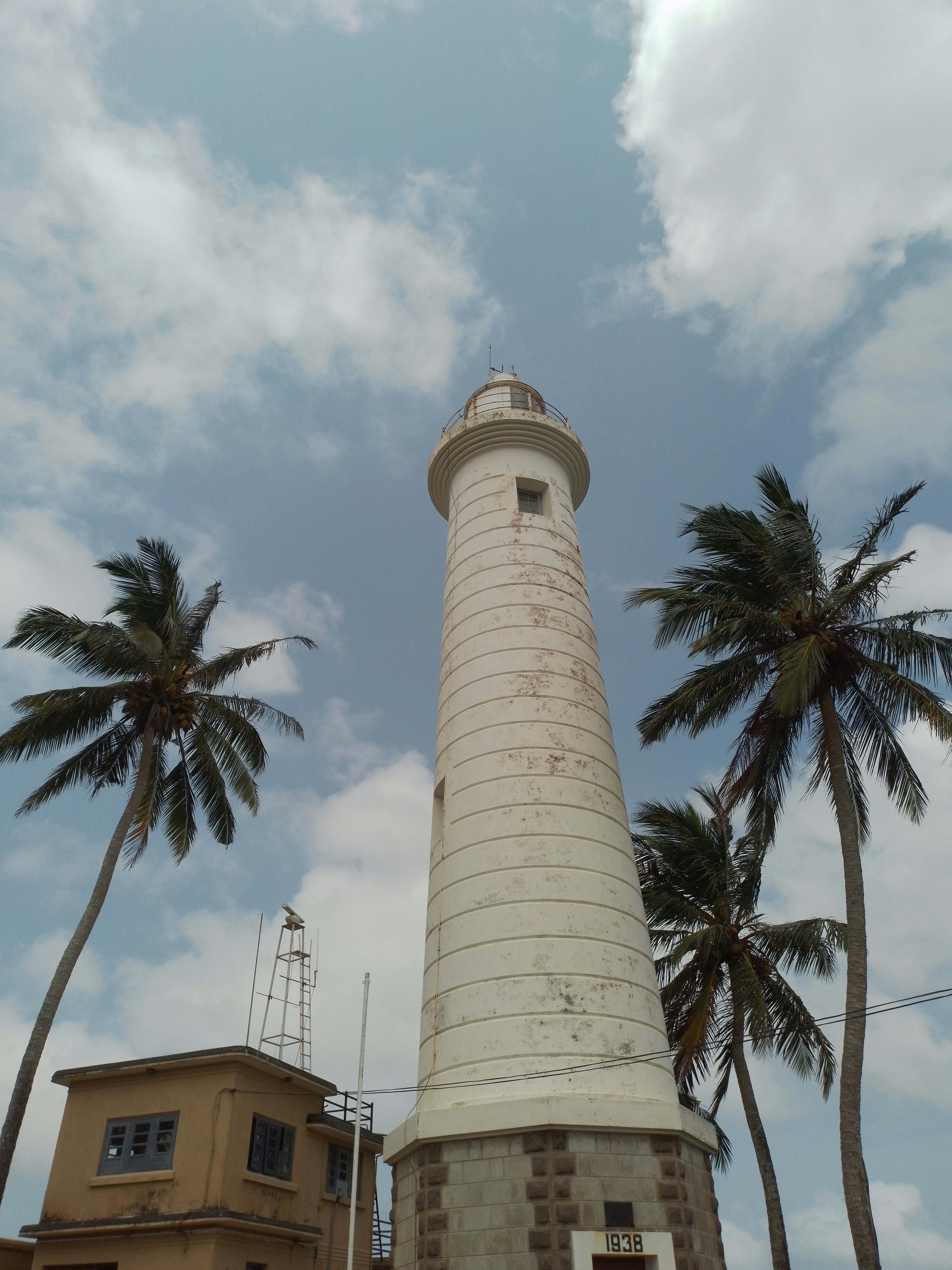 A tall white lighthouse rises between palm trees beneath a bright, cloud-dotted sky, creating a classic coastal beacon scene.