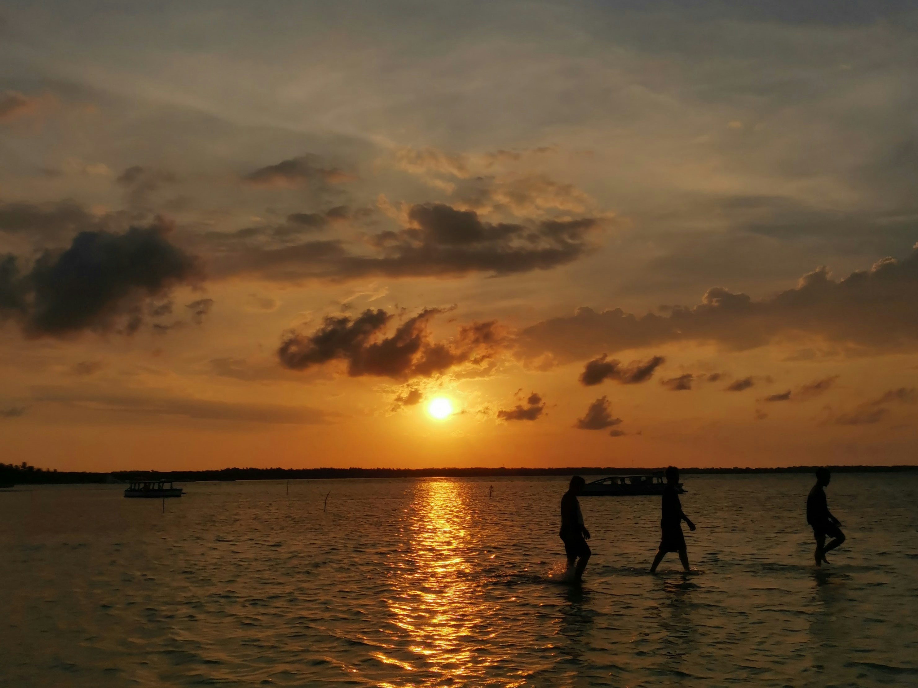 Silhouetted figures walk through shallow water as the sun sets, casting a warm glow across the sky and sea.