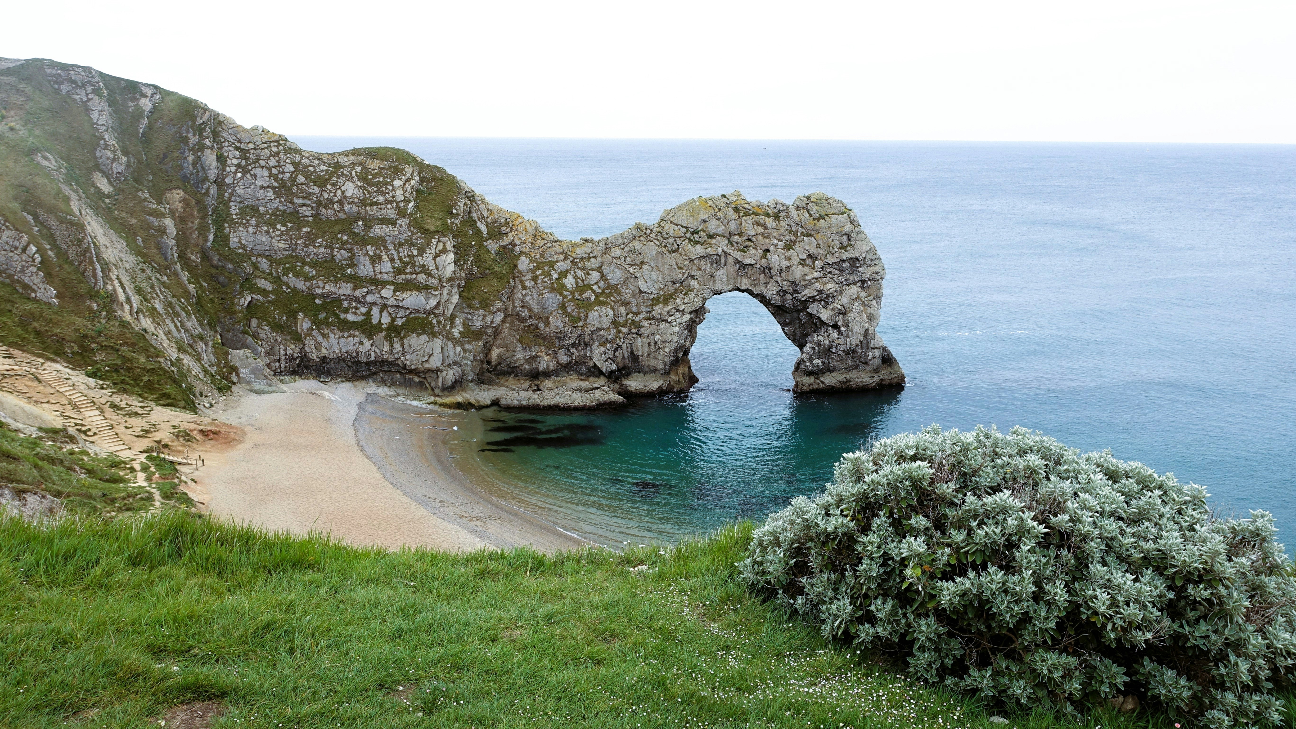 Rock arch rising from the sea near a sandy beach, framed by cliffs and greenery.