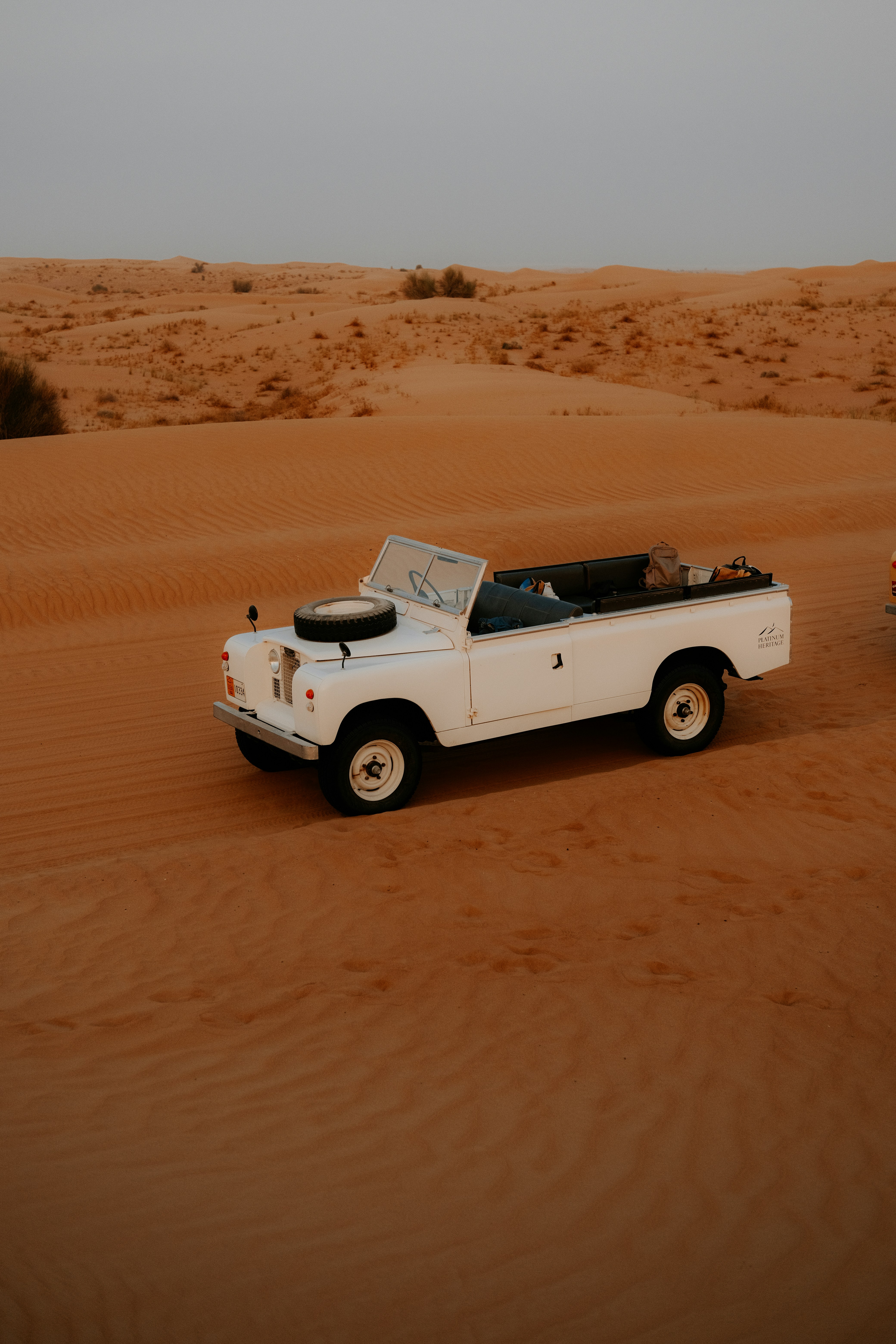 White vintage 4x4 parked on wind-sculpted orange dunes with a spare tire on the hood and gear in the back.