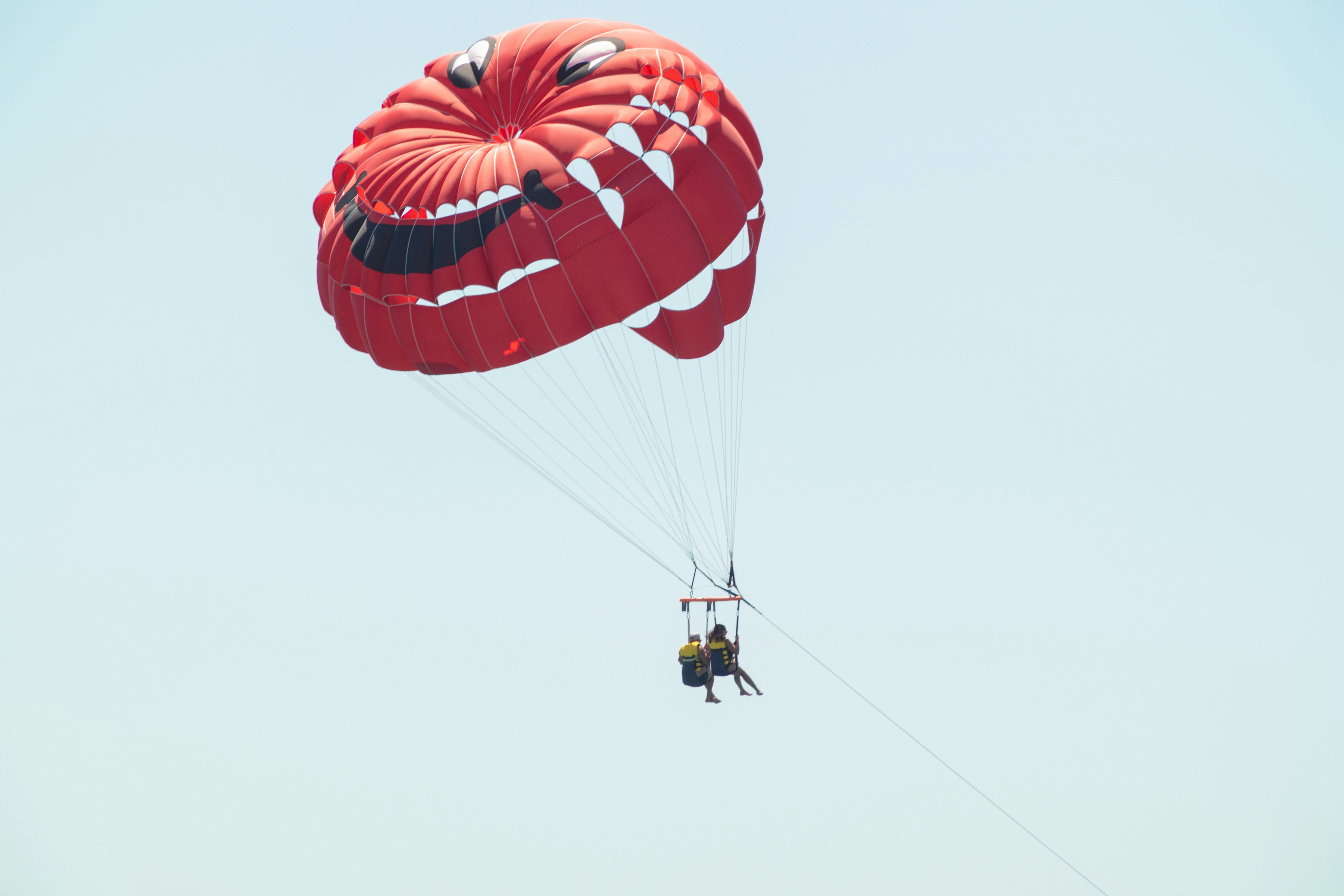 Two paragliders ride a red parachute against a pale blue sky, suspended by lines and a single guiding rope behind them. The scene highlights a moment of ascent and balance with minimal background distractions.