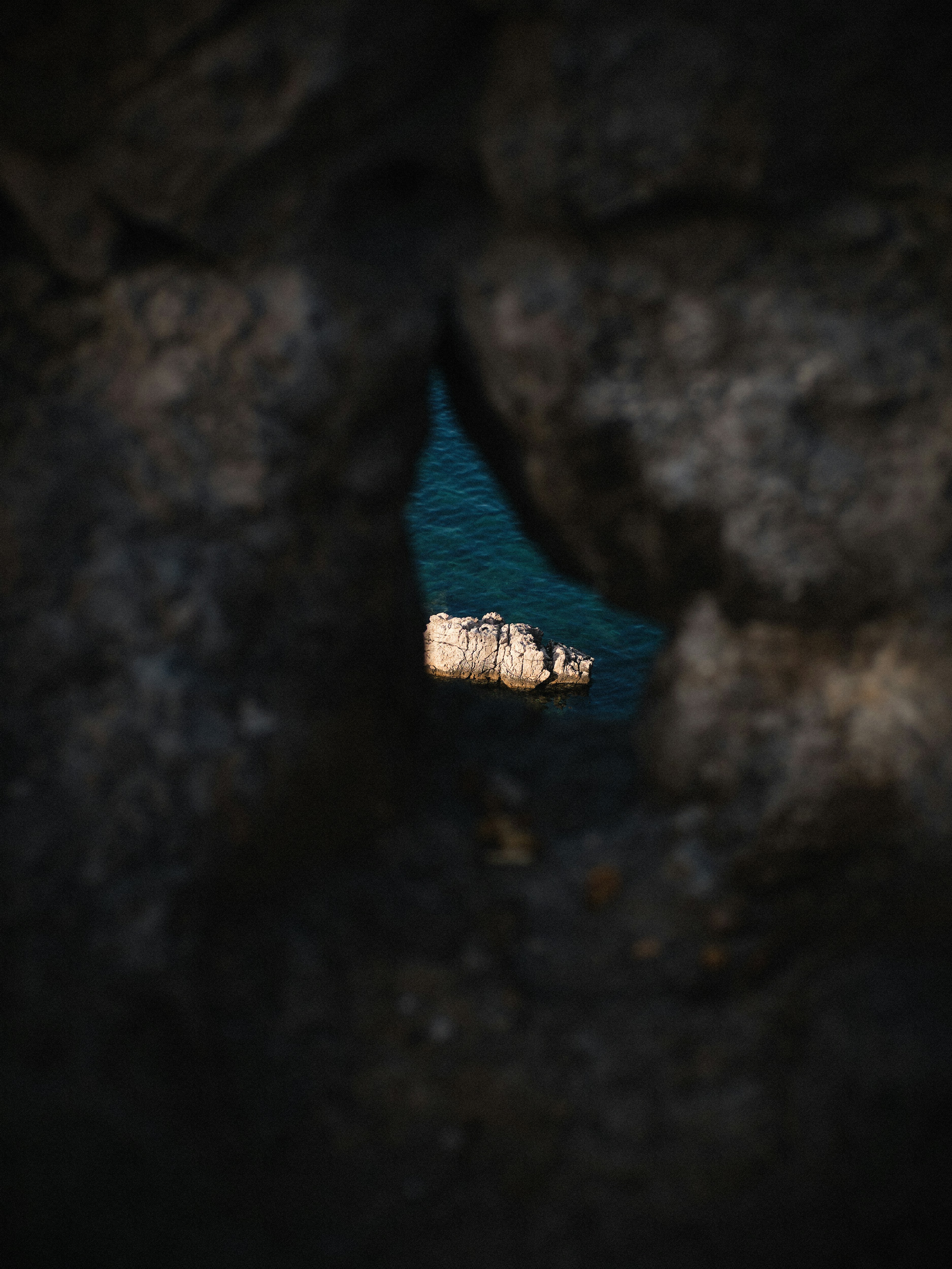 A narrow rocky window frames a sunlit rock island against turquoise water, while the dark, textured foreground dominates the scene.