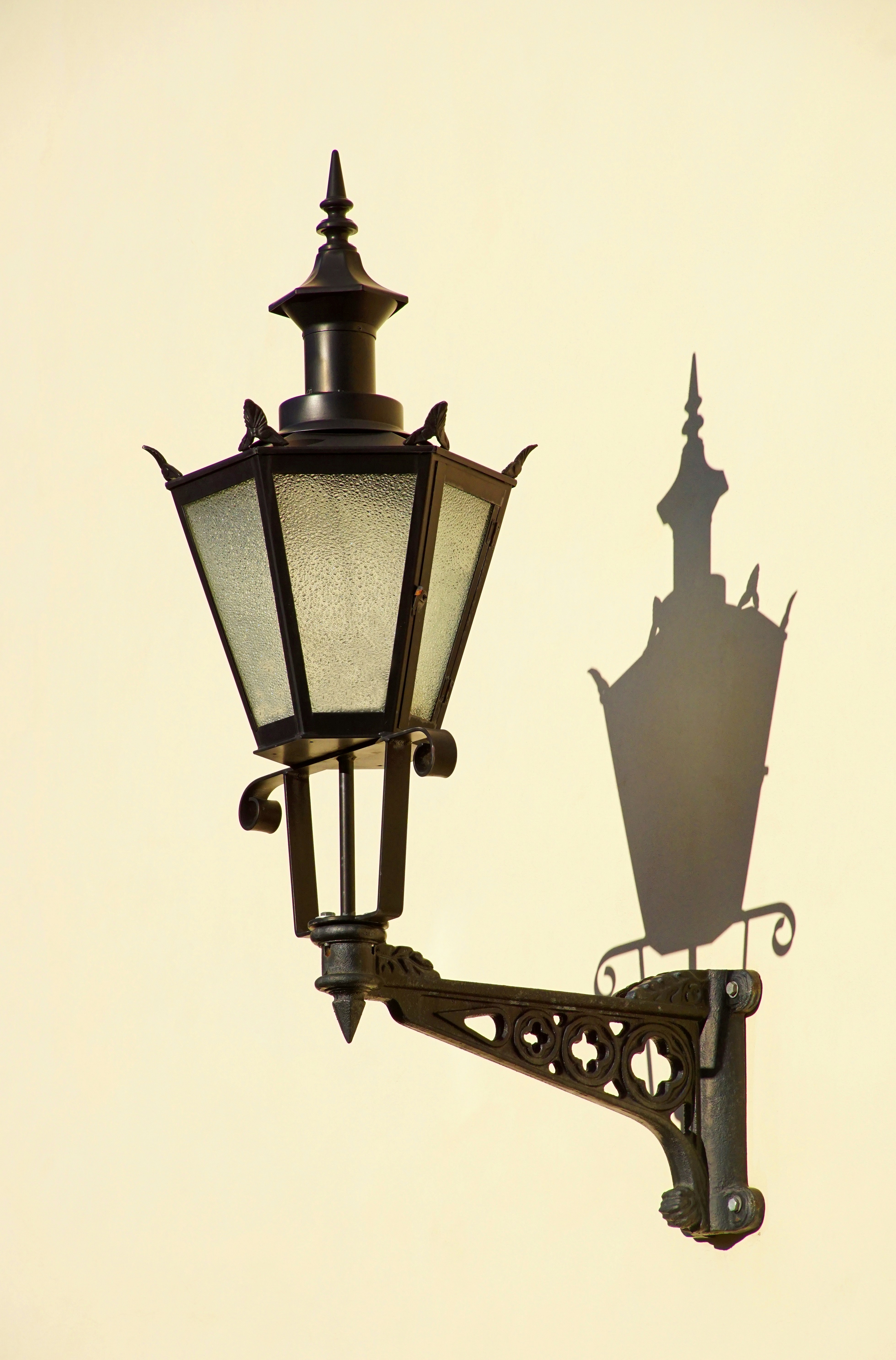 Photograph of an ornate street lantern mounted on a wrought-iron bracket against a pale cream wall, with its shadow stretching to the right.