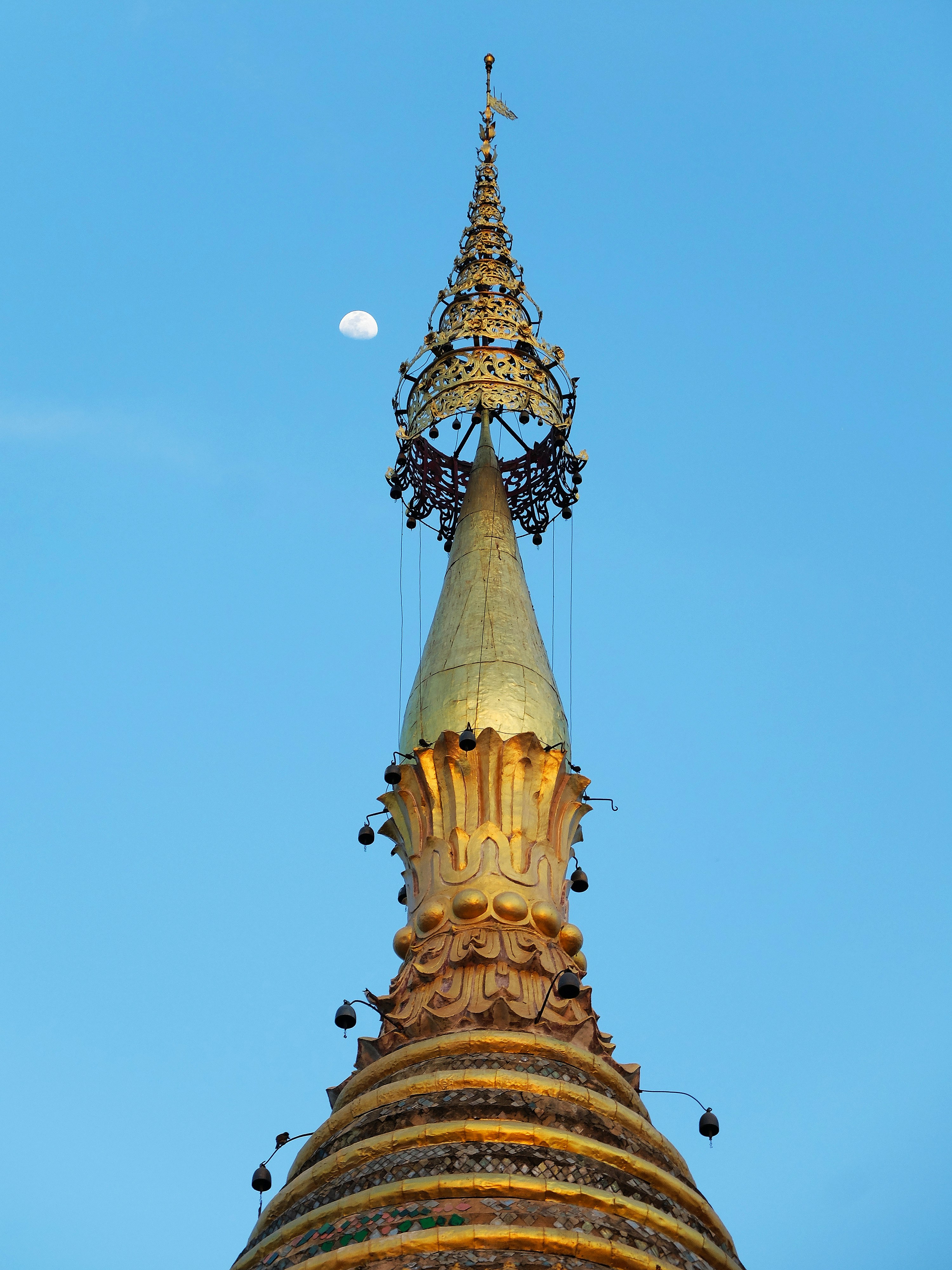 Golden spire of a temple reaching towards a clear blue sky, with the moon softly glowing in the background.