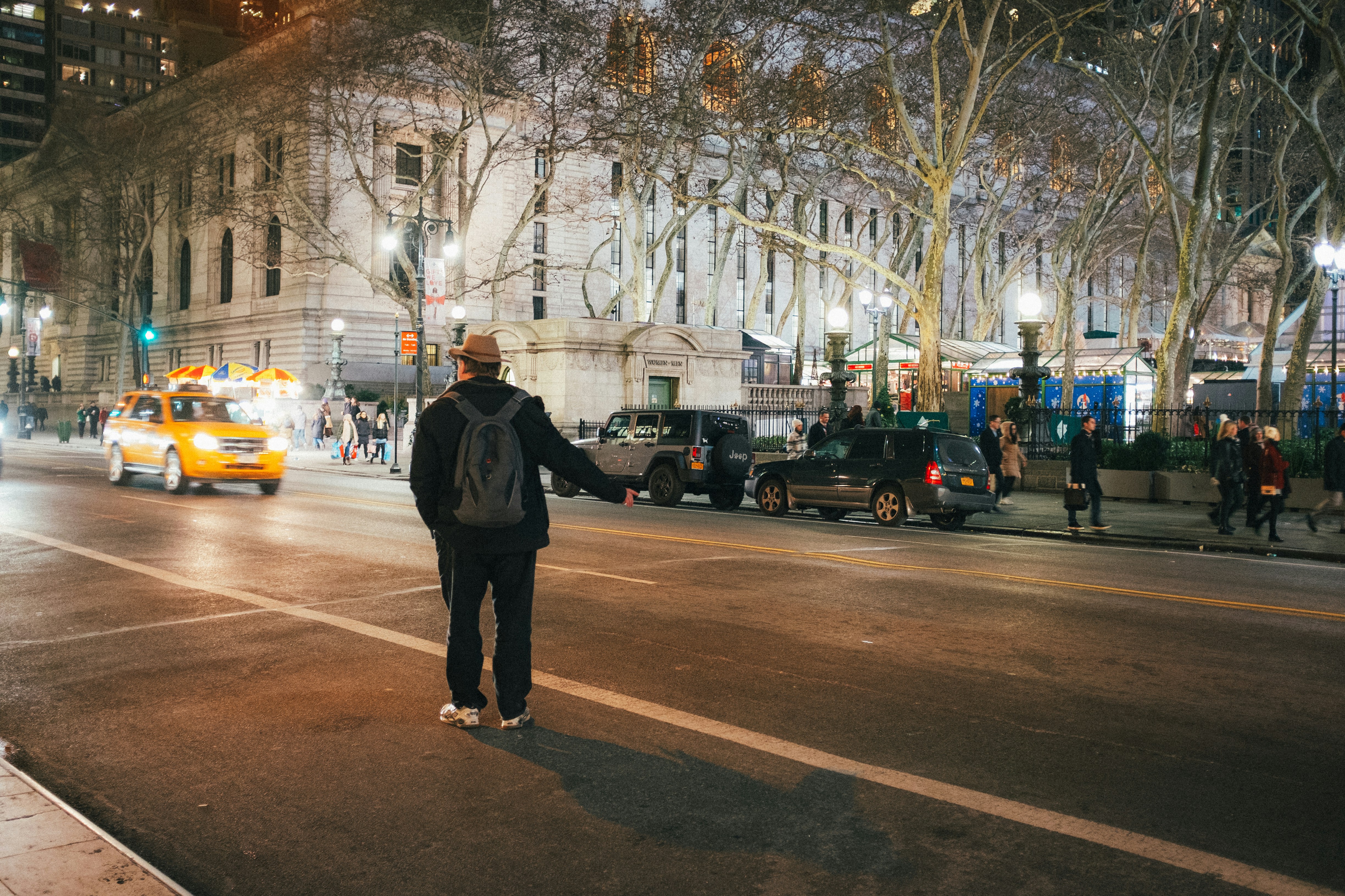Person standing on city street at night, signaling for an approaching yellow taxi.