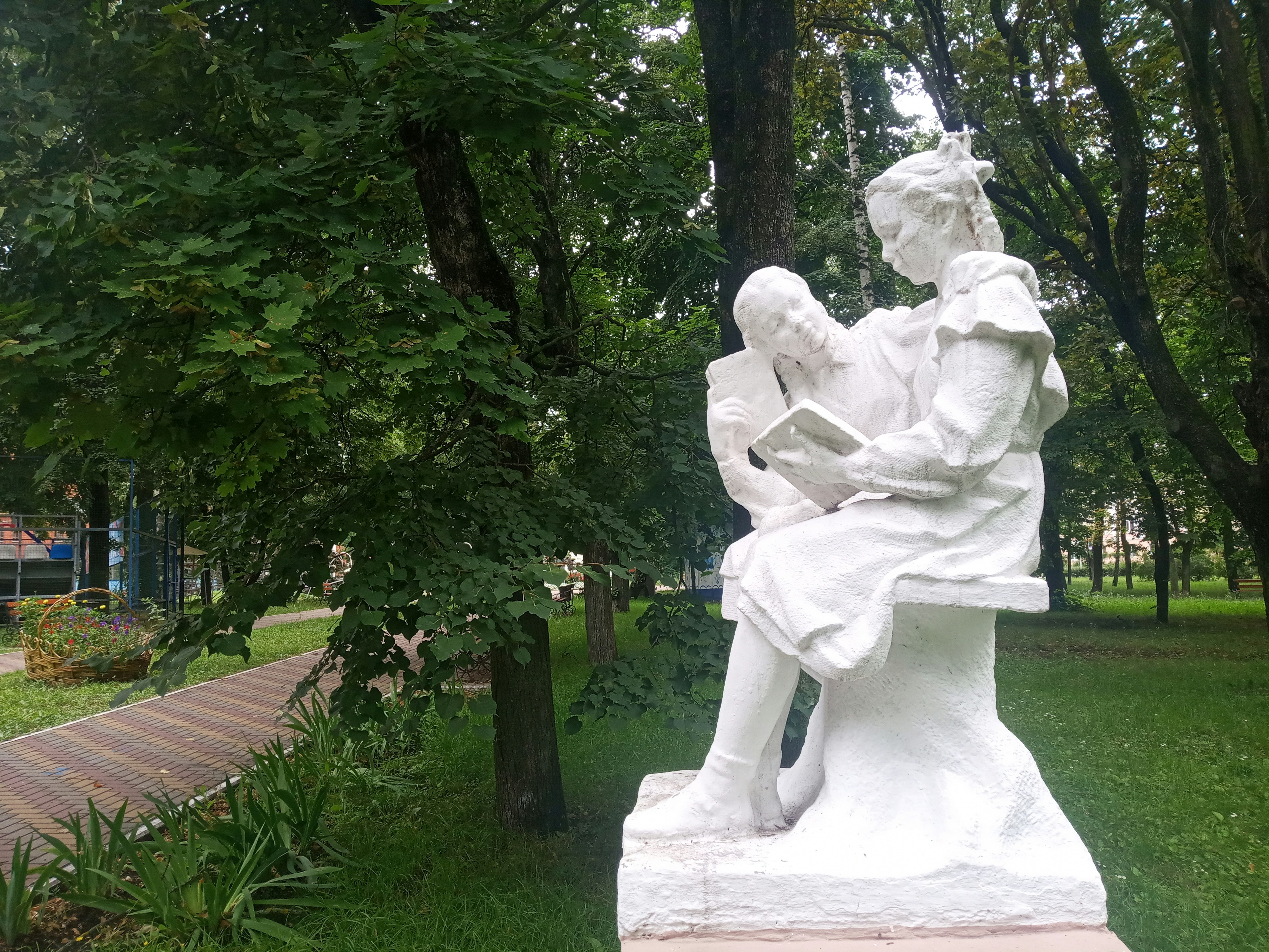 White marble statue of two figures reading a book in a green park, with a paved path and surrounding trees.