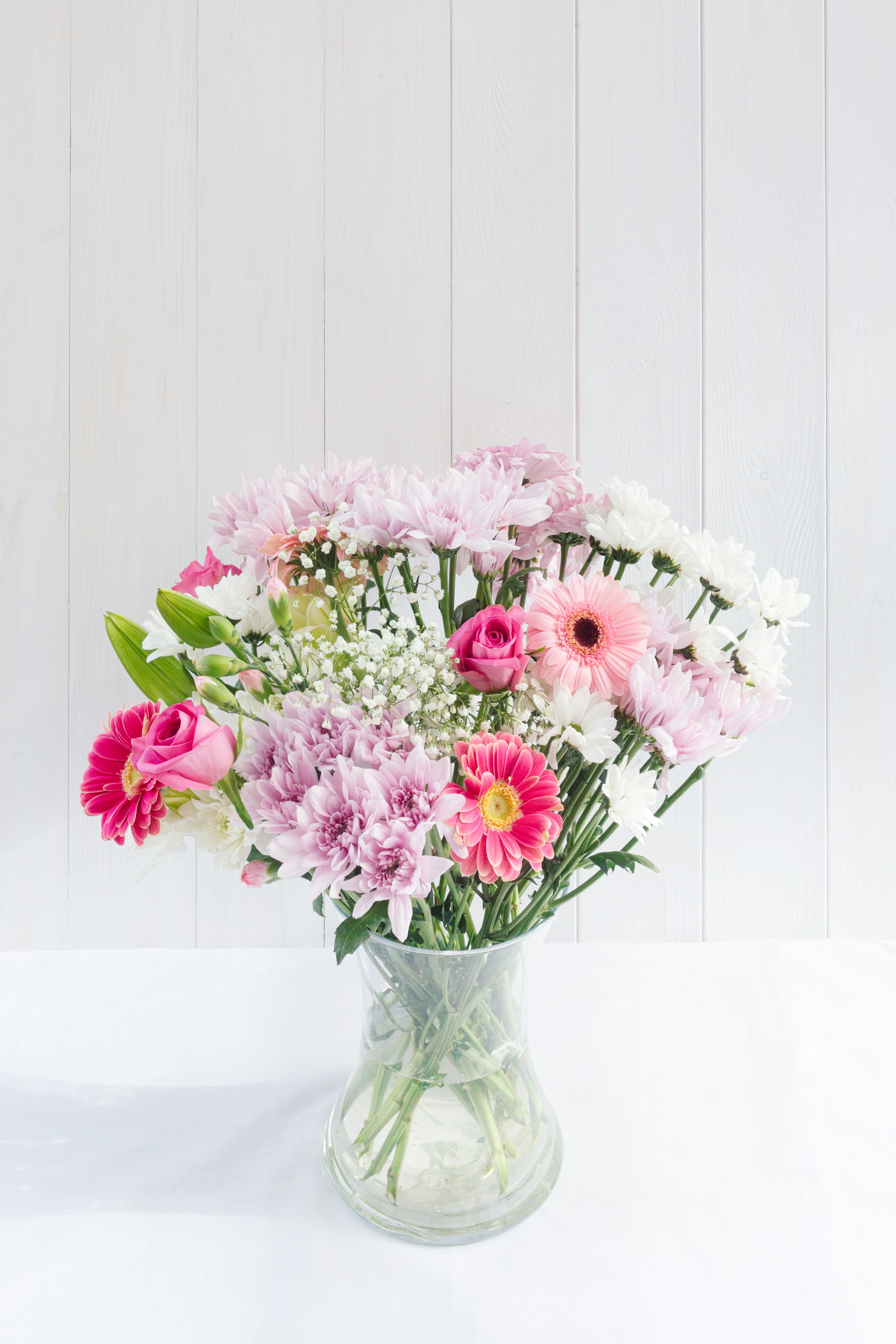 This is a colour photographic image of a pretty bunch of mixed flowers in a glass vase, set against a light background. There is some negative space above. The main colours of the image are pink, white and green.
