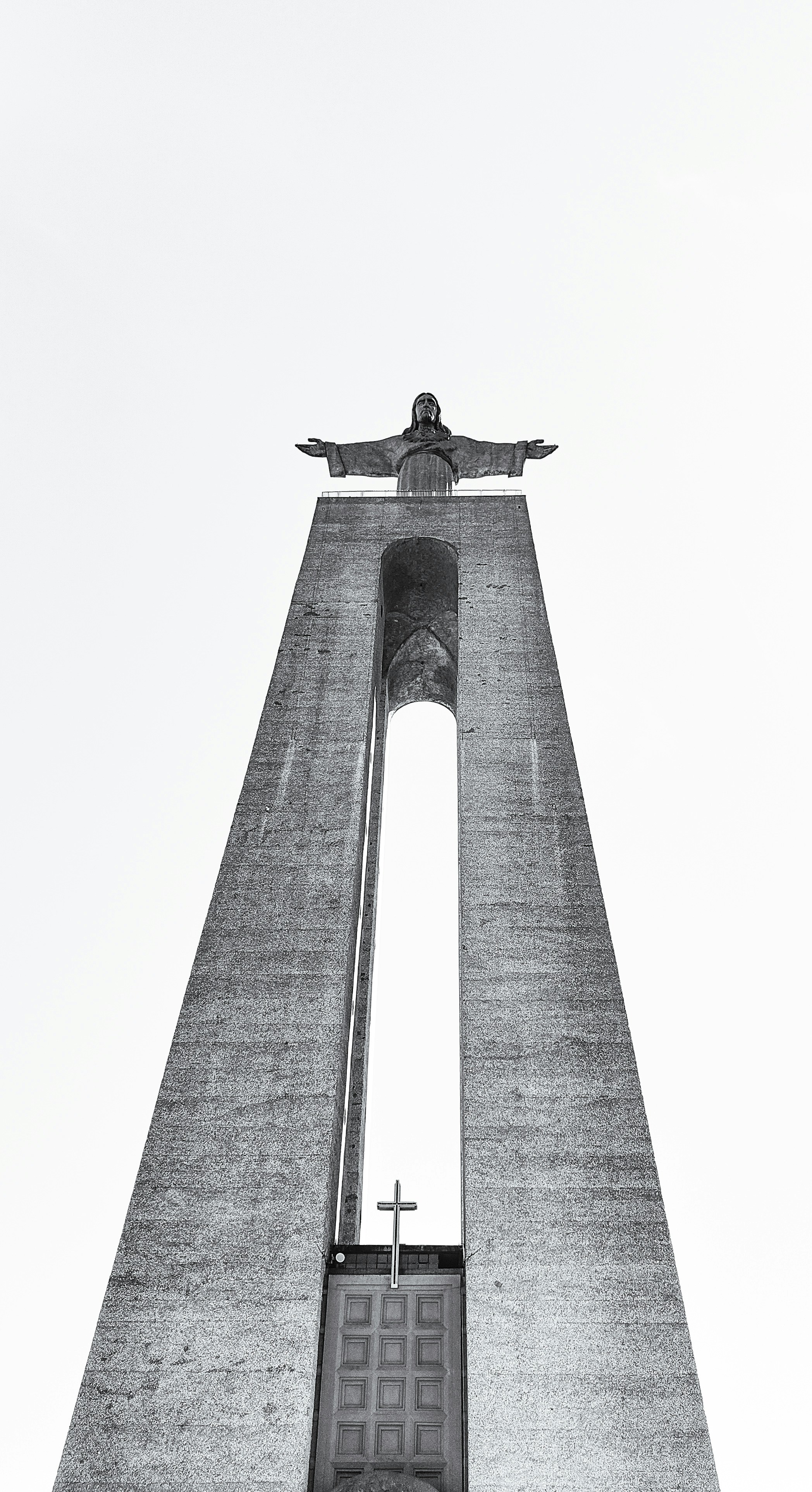 Photograph of a monumental statue with outstretched arms atop a tall arch, captured in high-contrast monochrome against a bright sky.