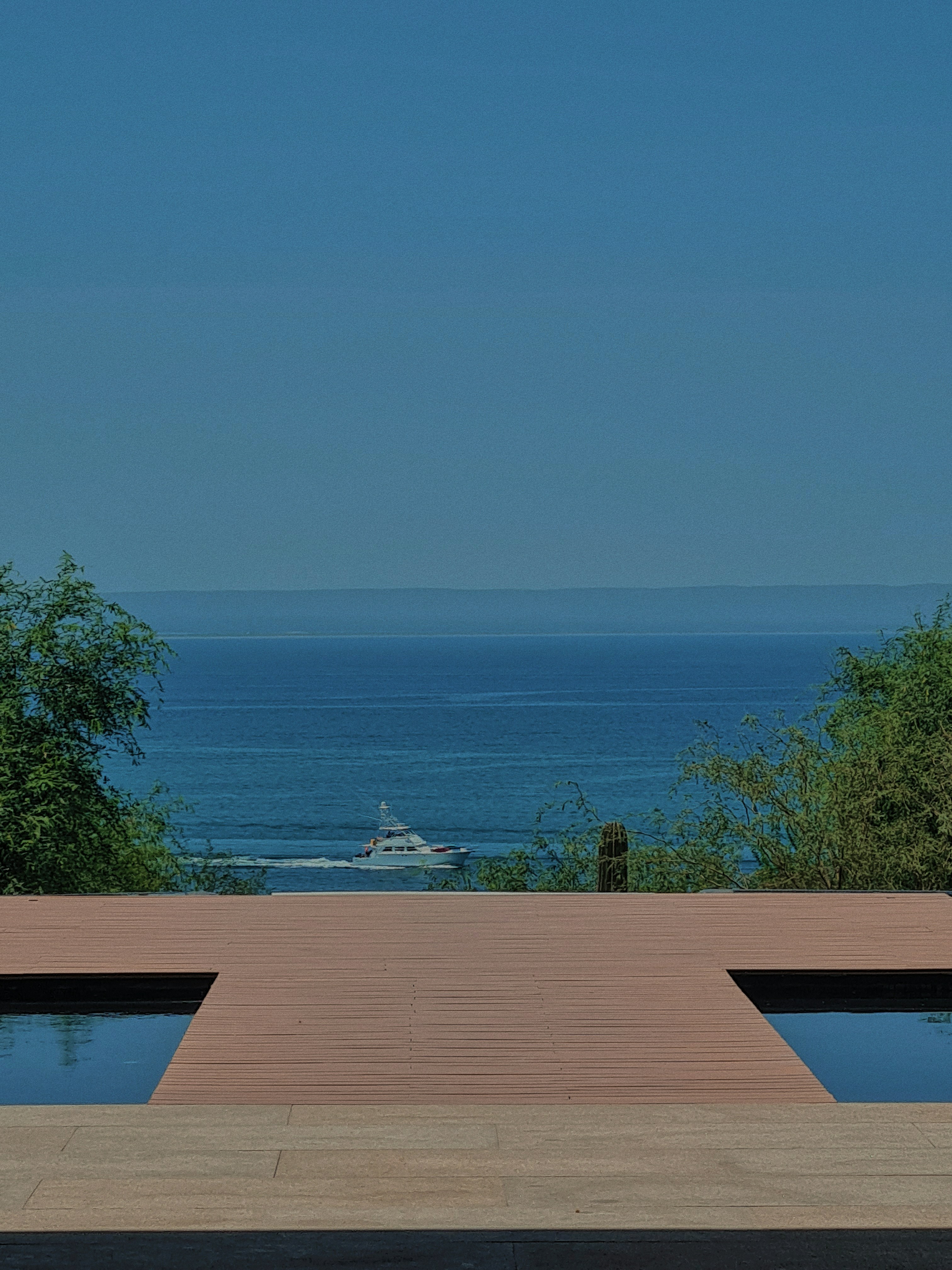 A modern terrace foreground frames a calm sea, with a small boat gliding along the horizon. Greenery on both sides guides the eye to the blue expanse beneath a clear sky.